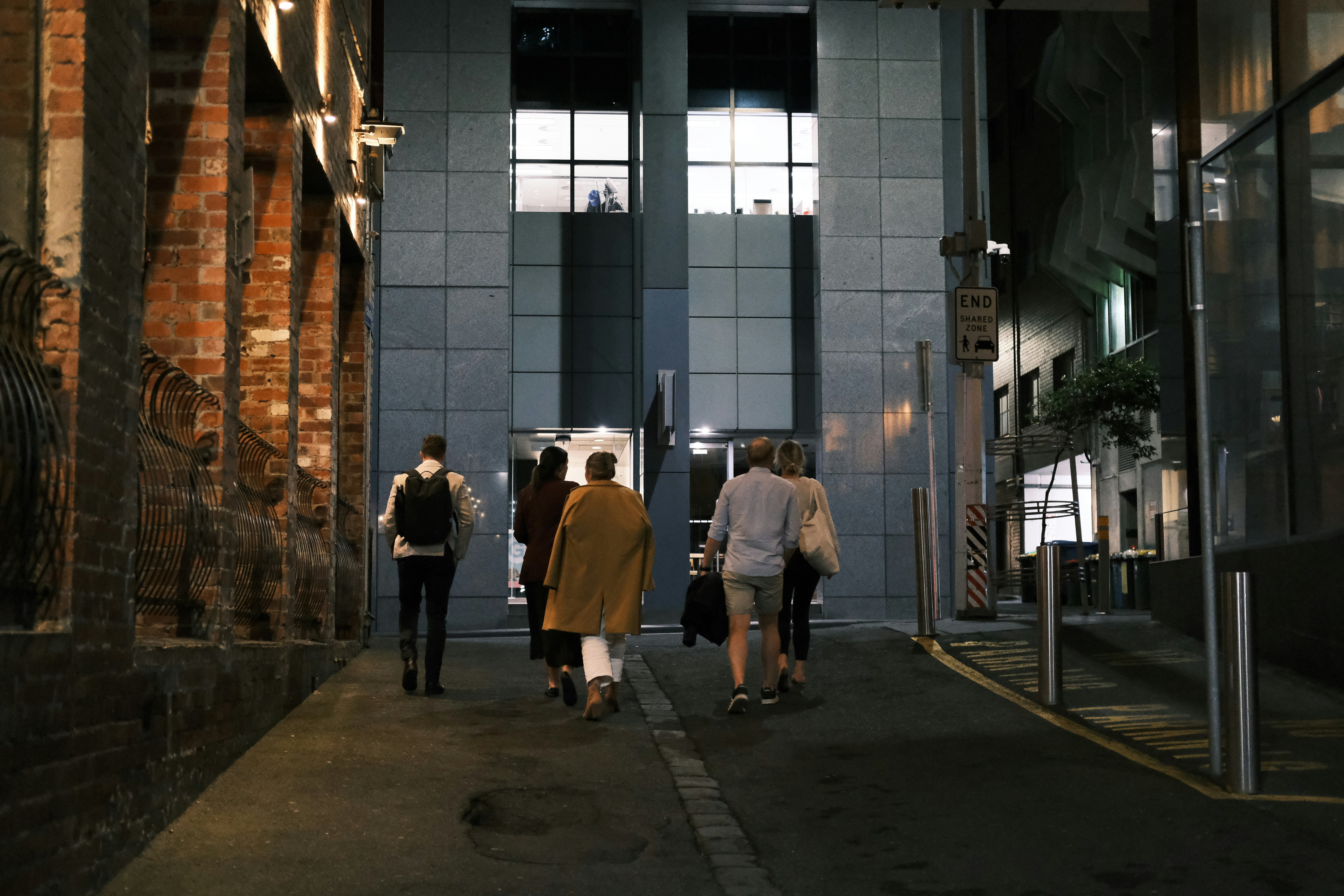 People walking through a dimly lit urban alley, flanked by brick and glass buildings with contrasting warm streetlights and cool evening tones.