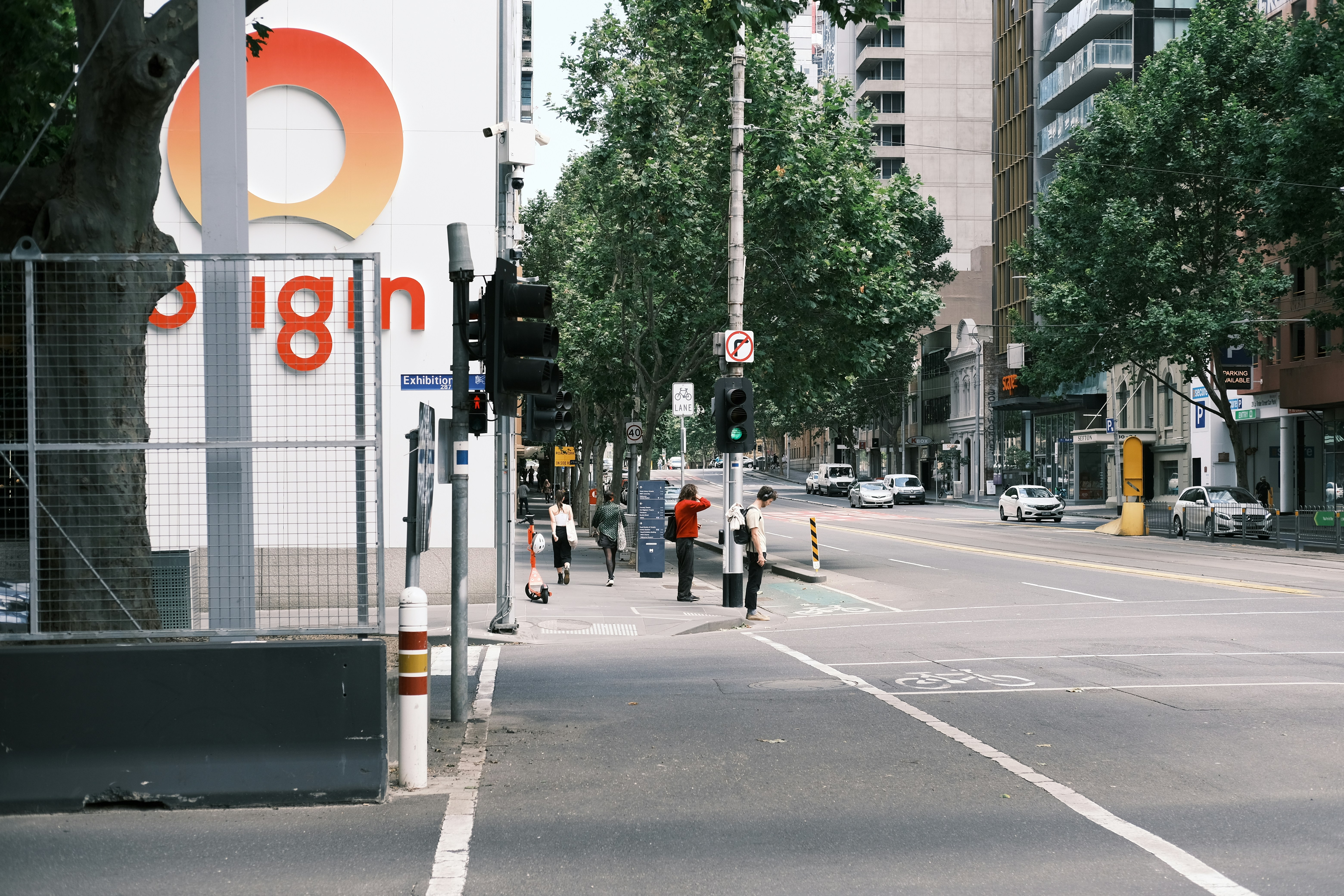 City intersection with people crossing under lush trees and tall buildings, featuring prominent signage.