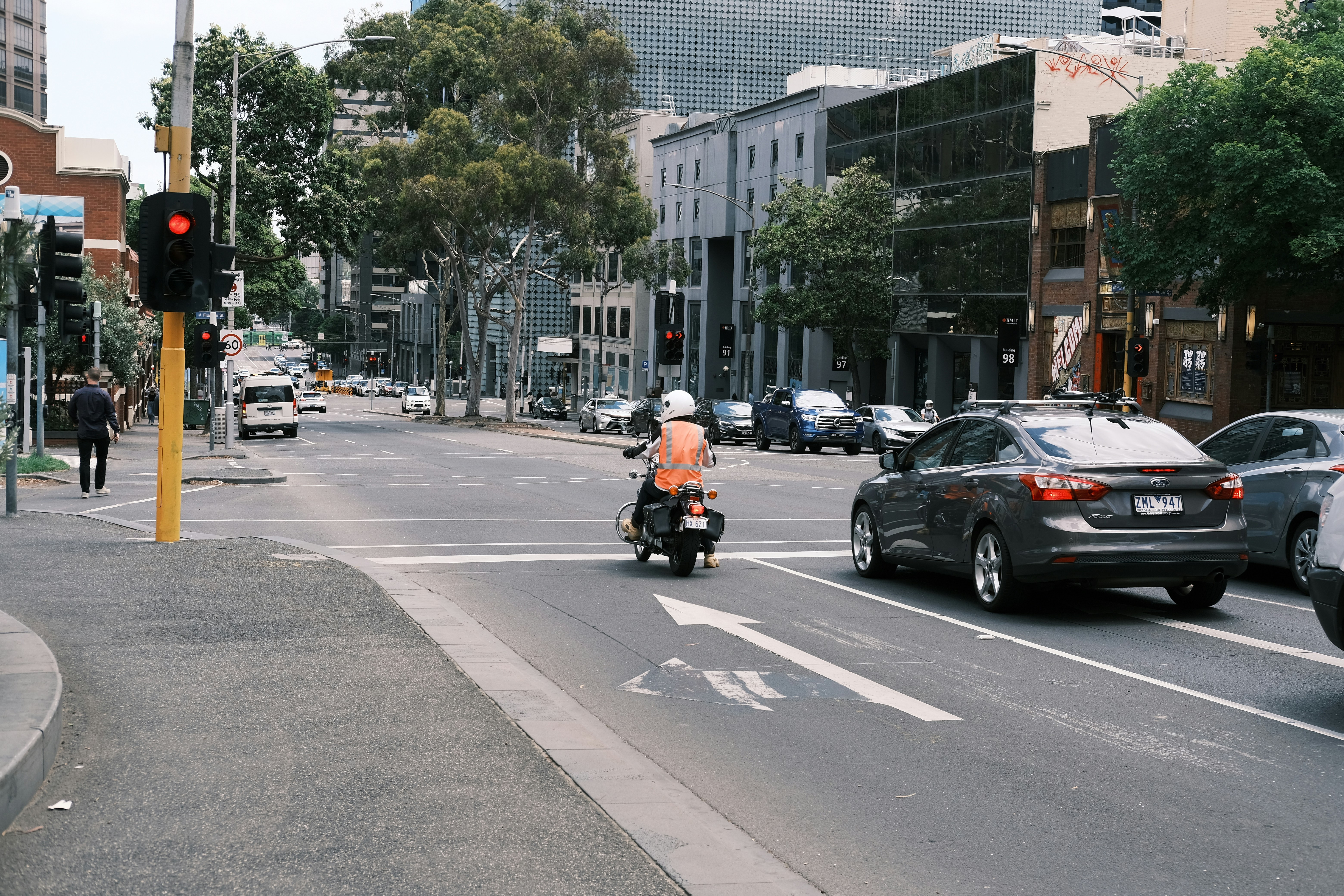 Motorcyclist in orange vest waits at a city intersection surrounded by modern buildings and trees under soft, overcast lighting.