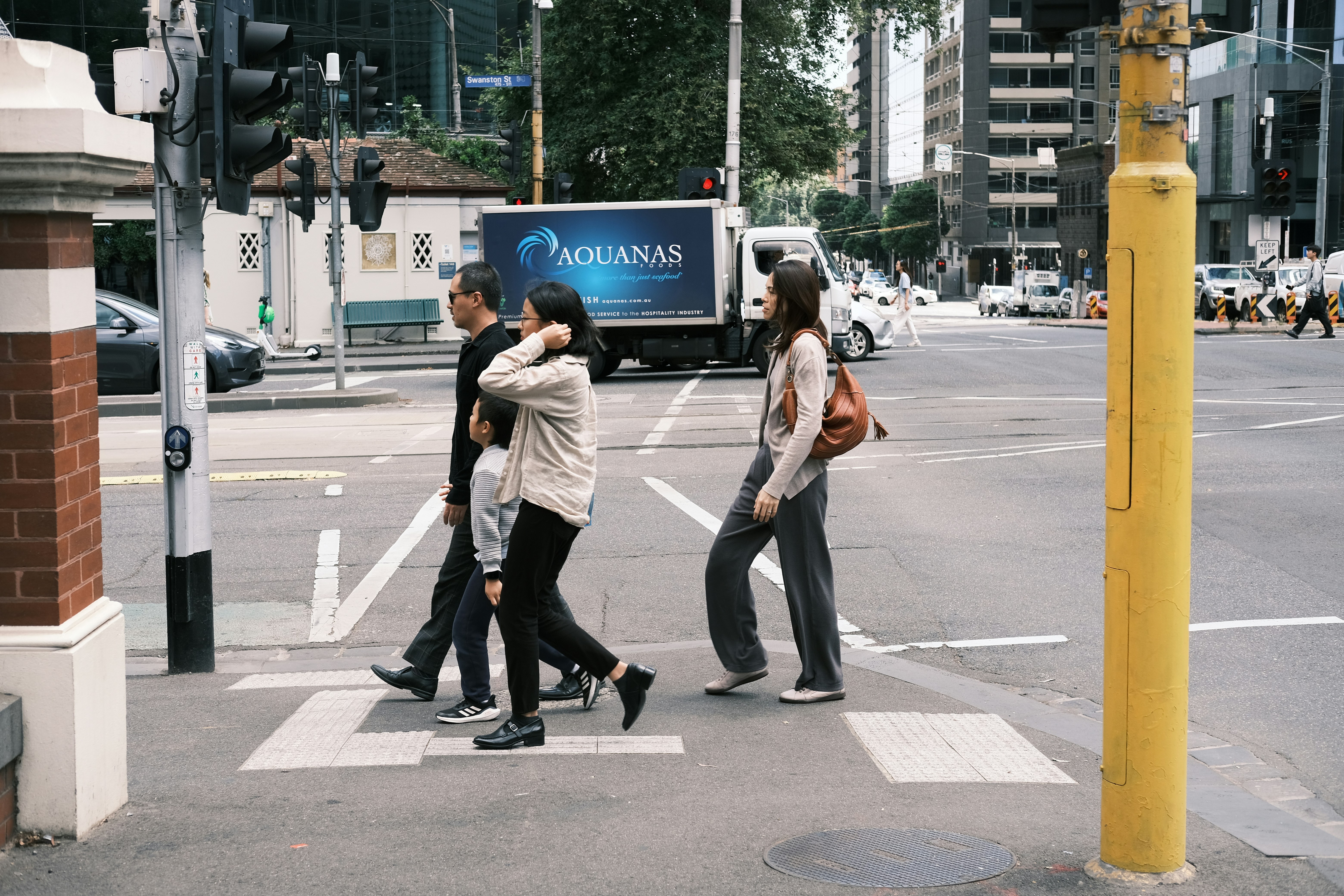 Pedestrians traverse a lively city crosswalk with modern architecture and vibrant urban elements, including a yellow traffic pole and blue truck advertisement.