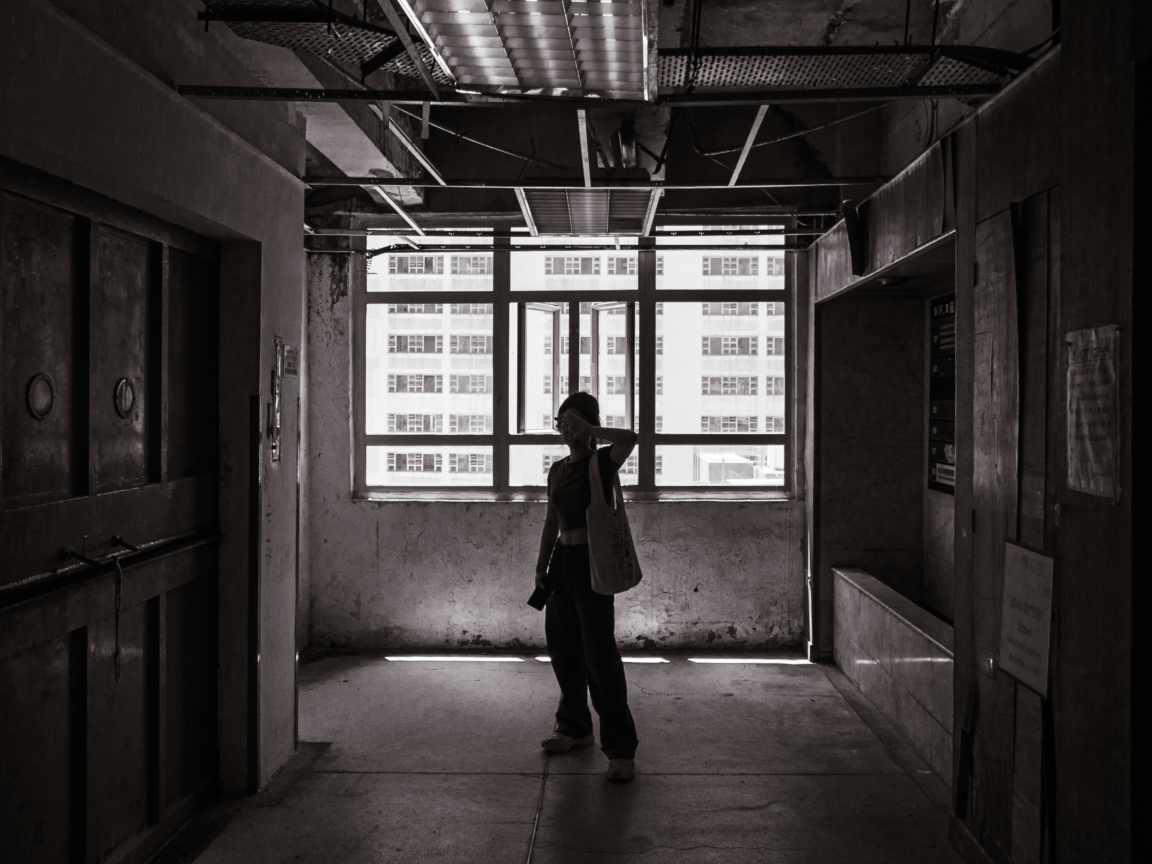 A lone figure stands in a dim, industrial hallway, backlit by large grid windows that reveal daylight outside.