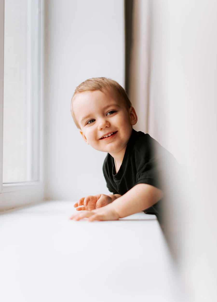 Young child leans on a white windowsill, smiling toward the camera in a softly lit indoor scene; a candid portrait with a shallow depth of field emphasizing the subject. Photo appears as a natural candid capture.