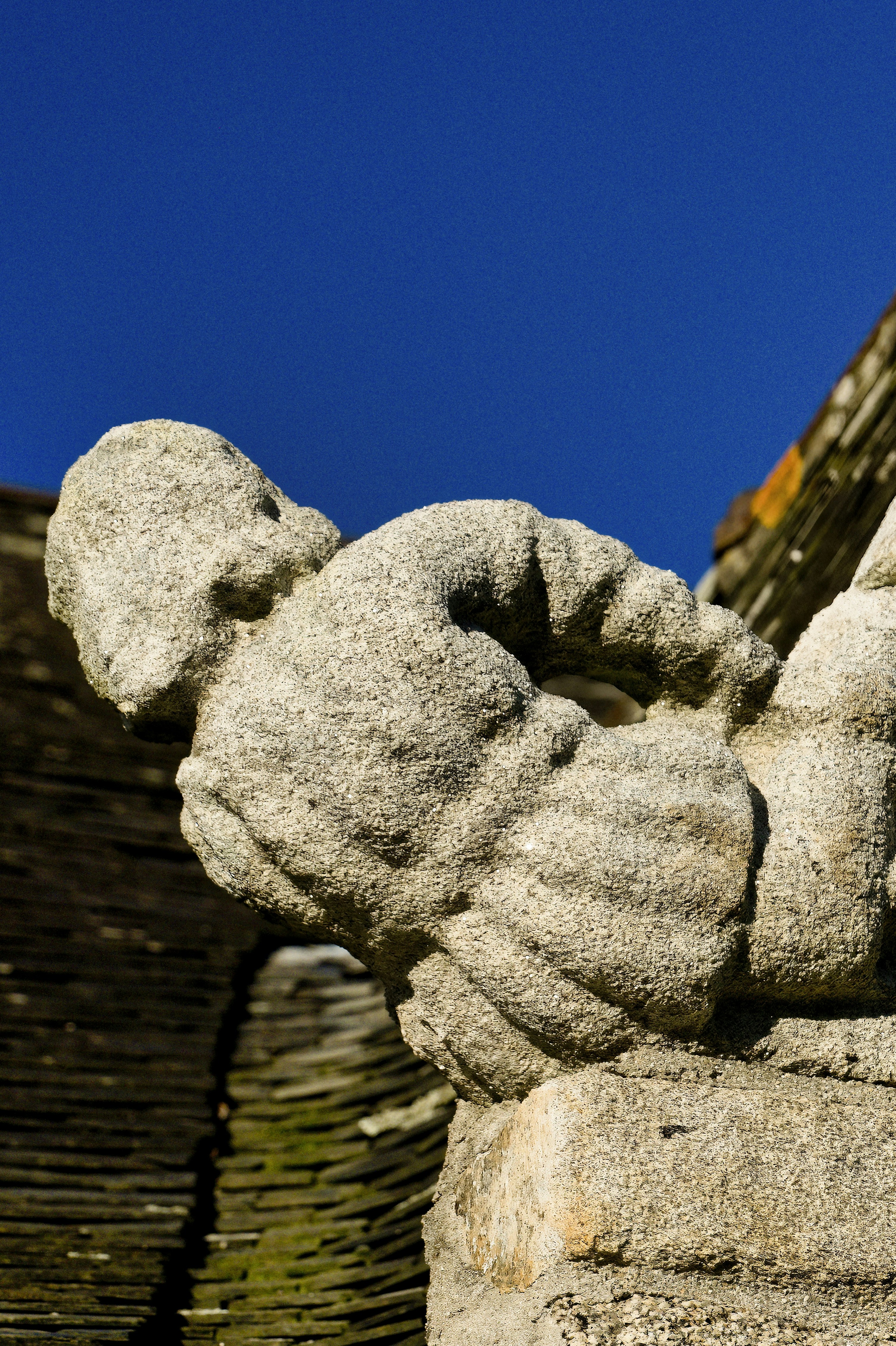 Sculpted figure resting against a rustic stone structure under a clear blue sky - A detailed stone sculpture of a reclining figure is positioned on the edge of an ancient building. The textures of the statue contrast with the smooth sky above.