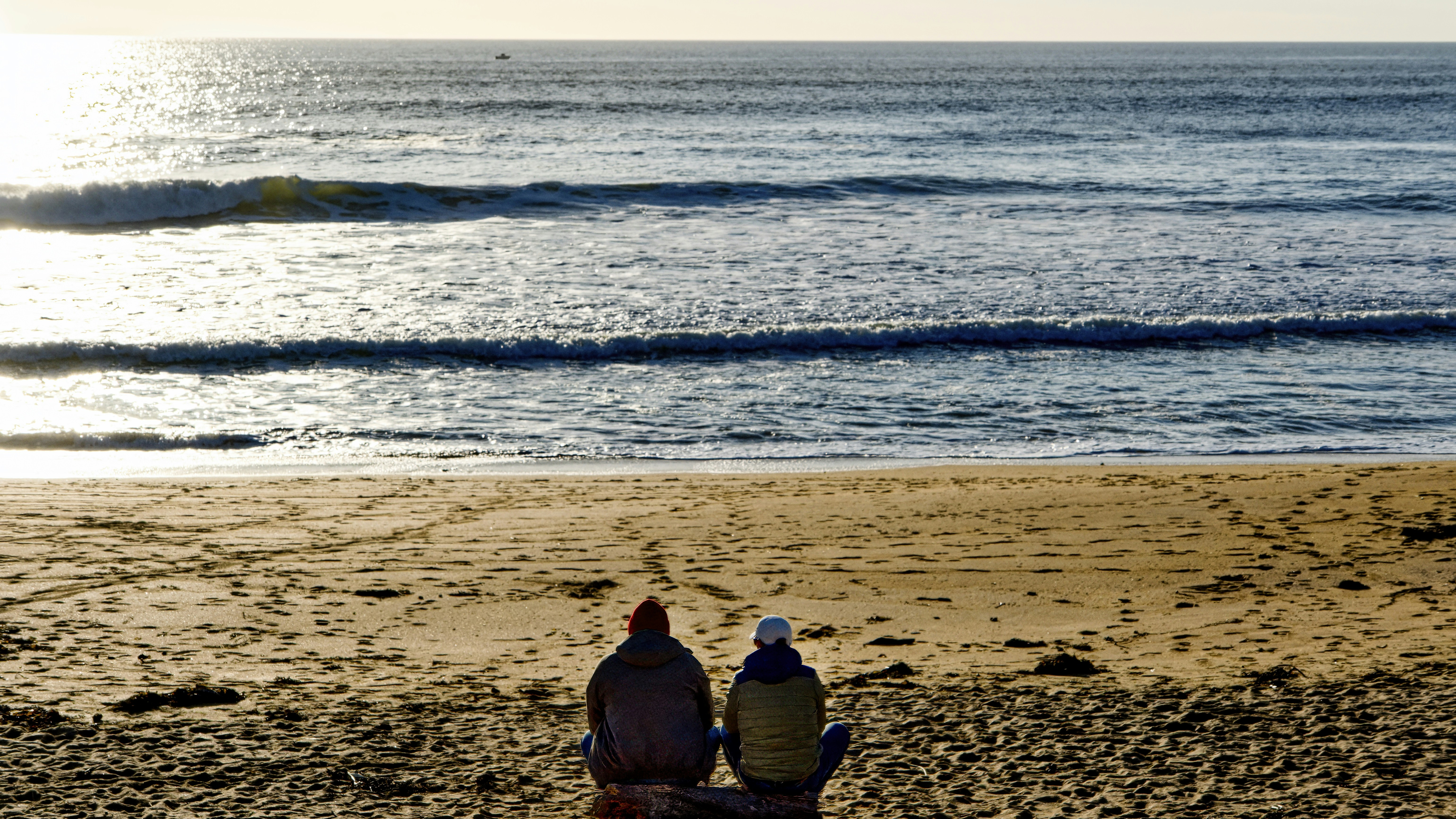 Couple seated on sandy beach, observing waves at sunset under a clear sky.