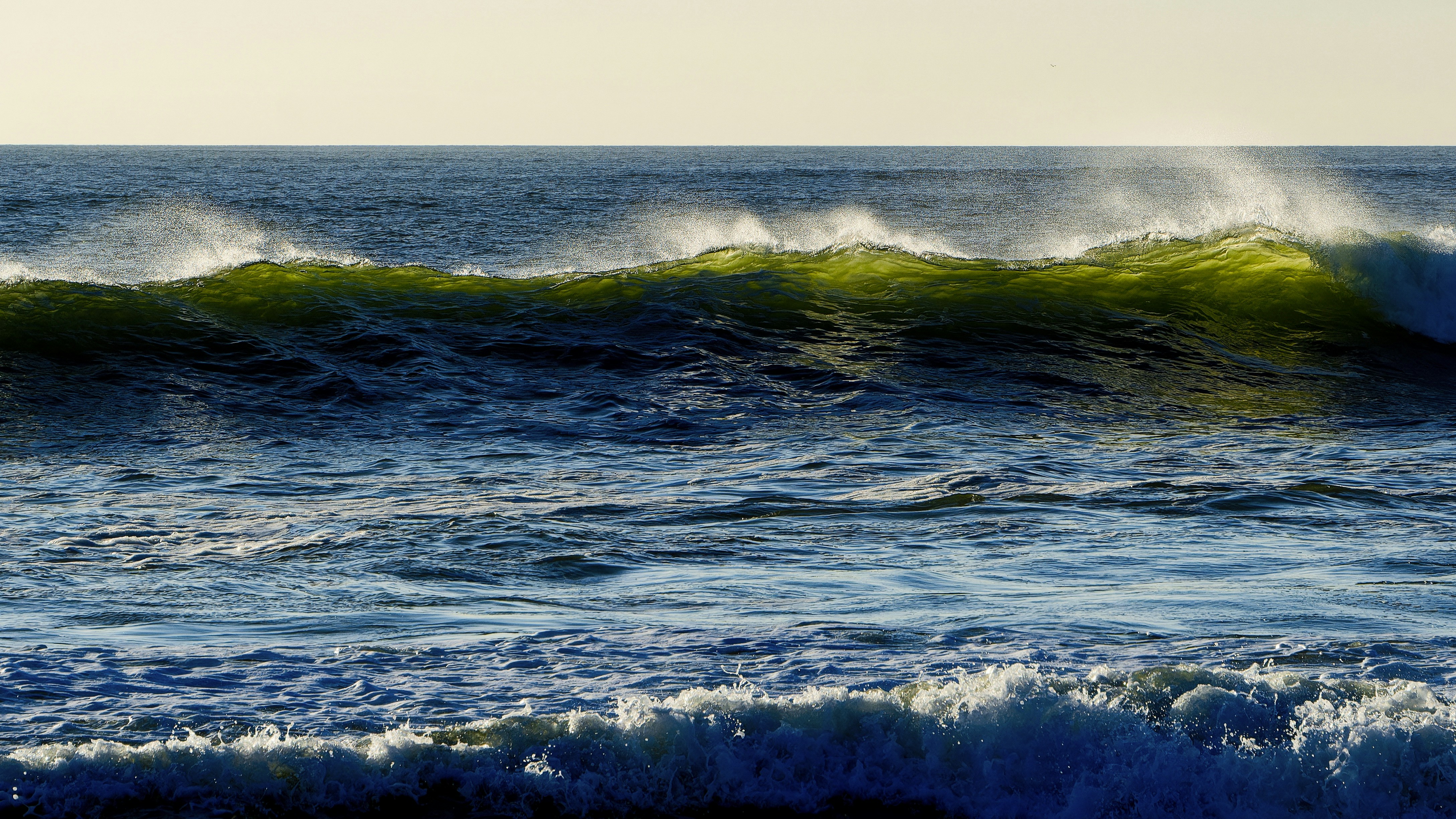 Dynamic ocean waves rolling towards the shore, illuminated by soft sunlight. The scene captures the vibrant hues of green and blue in the water.