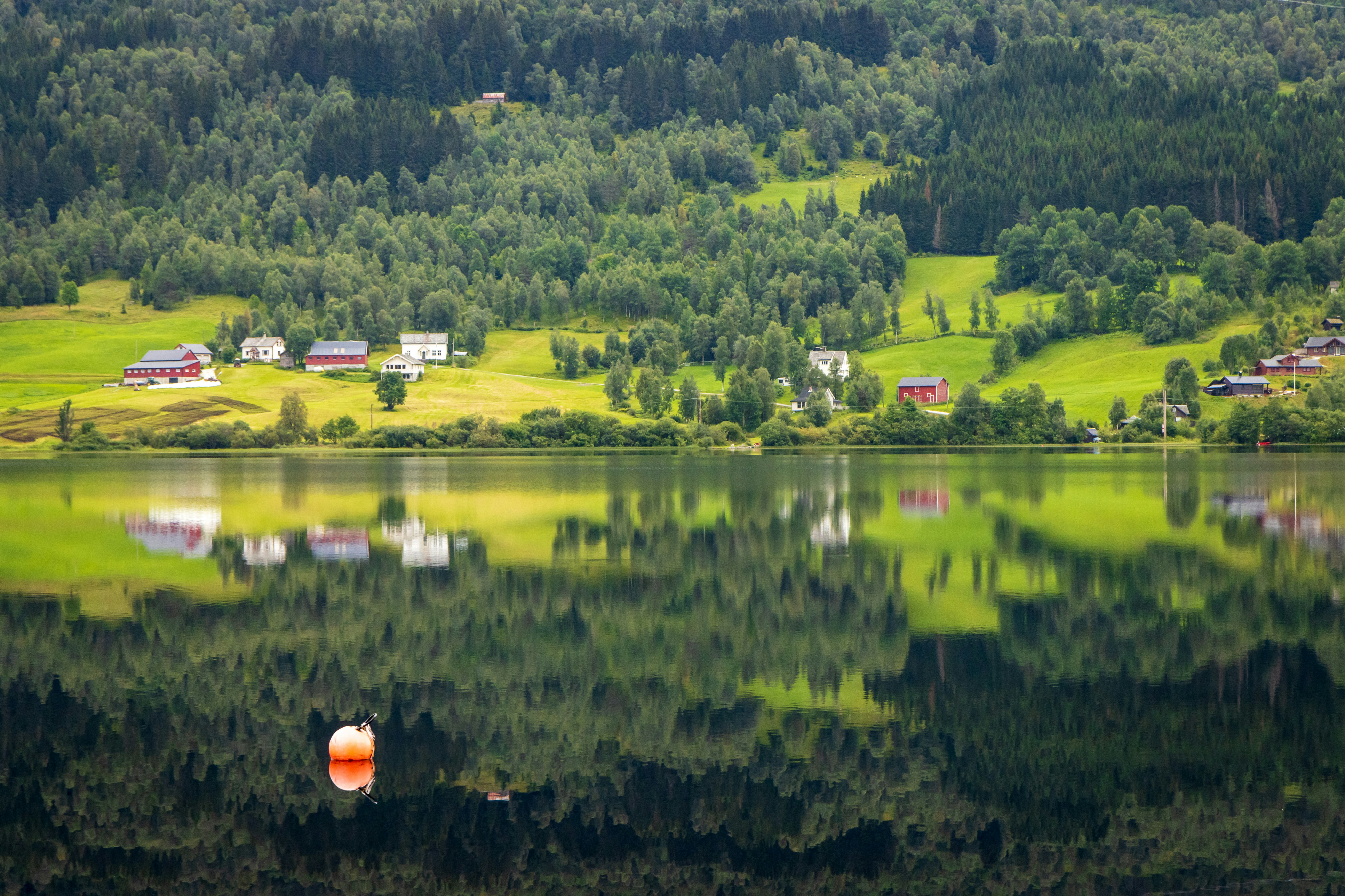 Lush green landscape reflected in a calm lake with red and white houses and an orange buoy as a focal point.