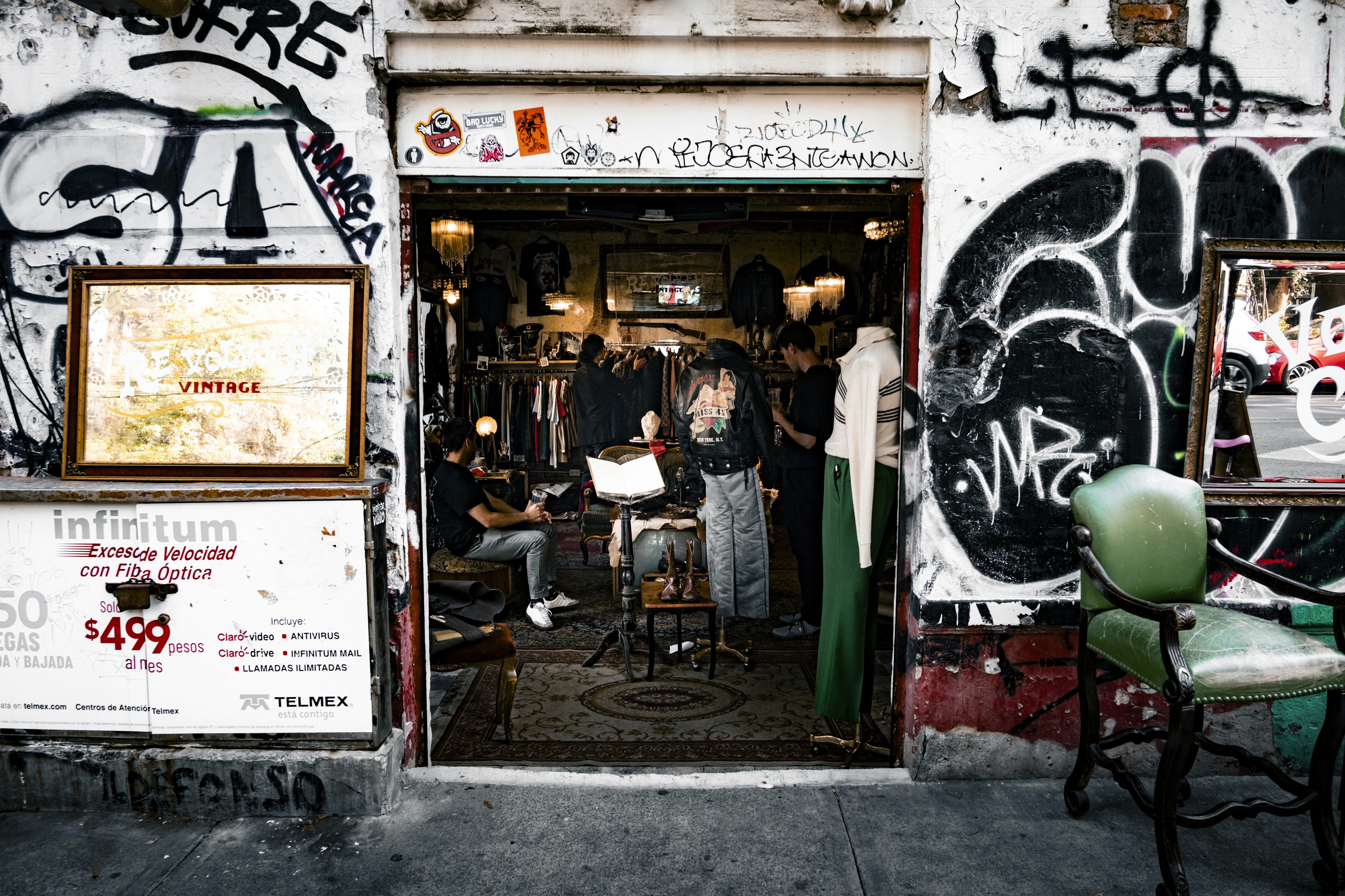 Entrance to a vintage boutique surrounded by graffiti, with warm chandeliers lighting retro clothing inside.