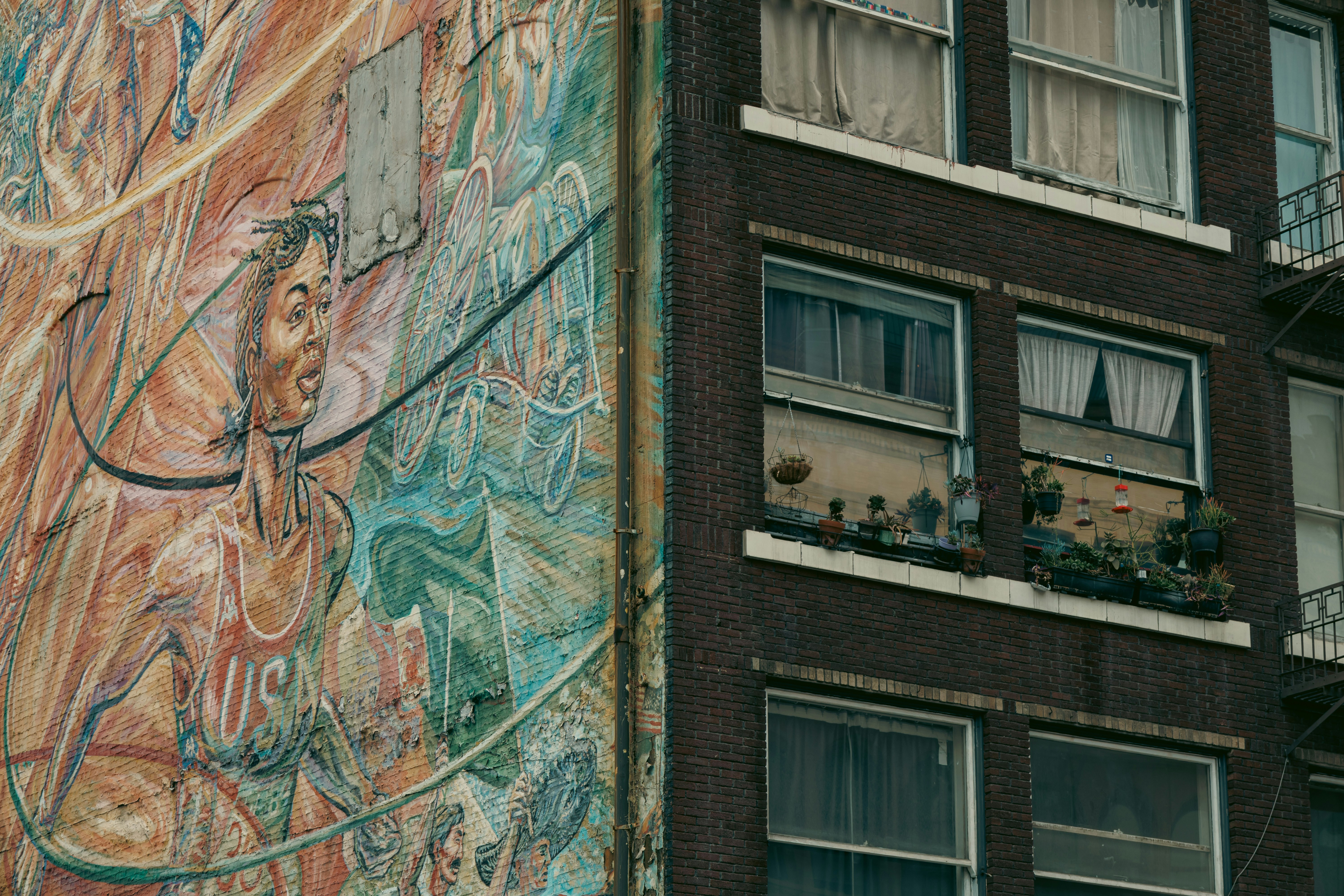 Colorful mural depicting athletes in motion beside a brick residential building with potted plants on windowsills.