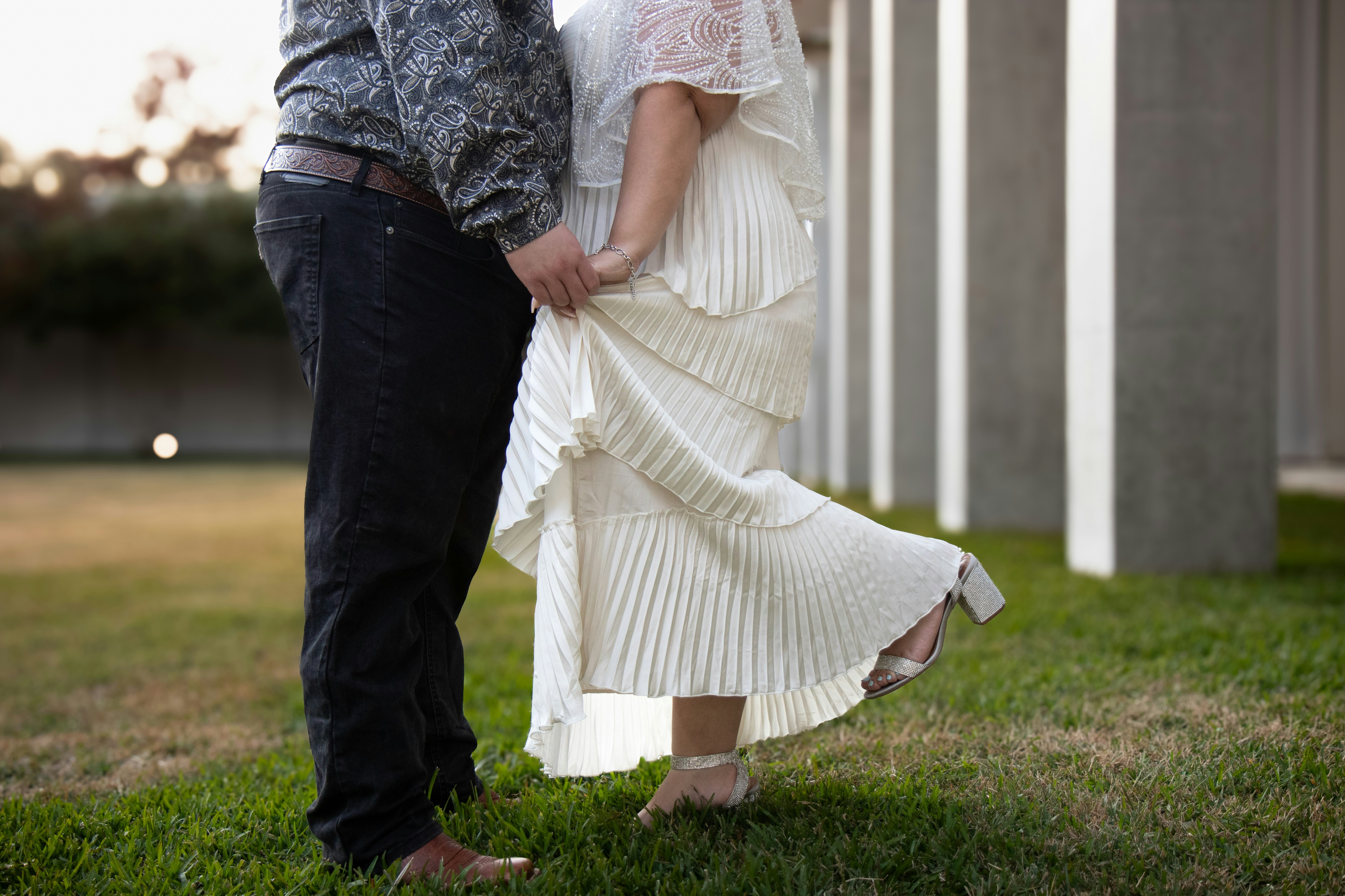 Couple holding hands with focus on their lower halves, standing in a tranquil outdoor setting. The woman's pleated white dress contrasts with the man's dark pants, enhanced by soft natural light and lush grass.