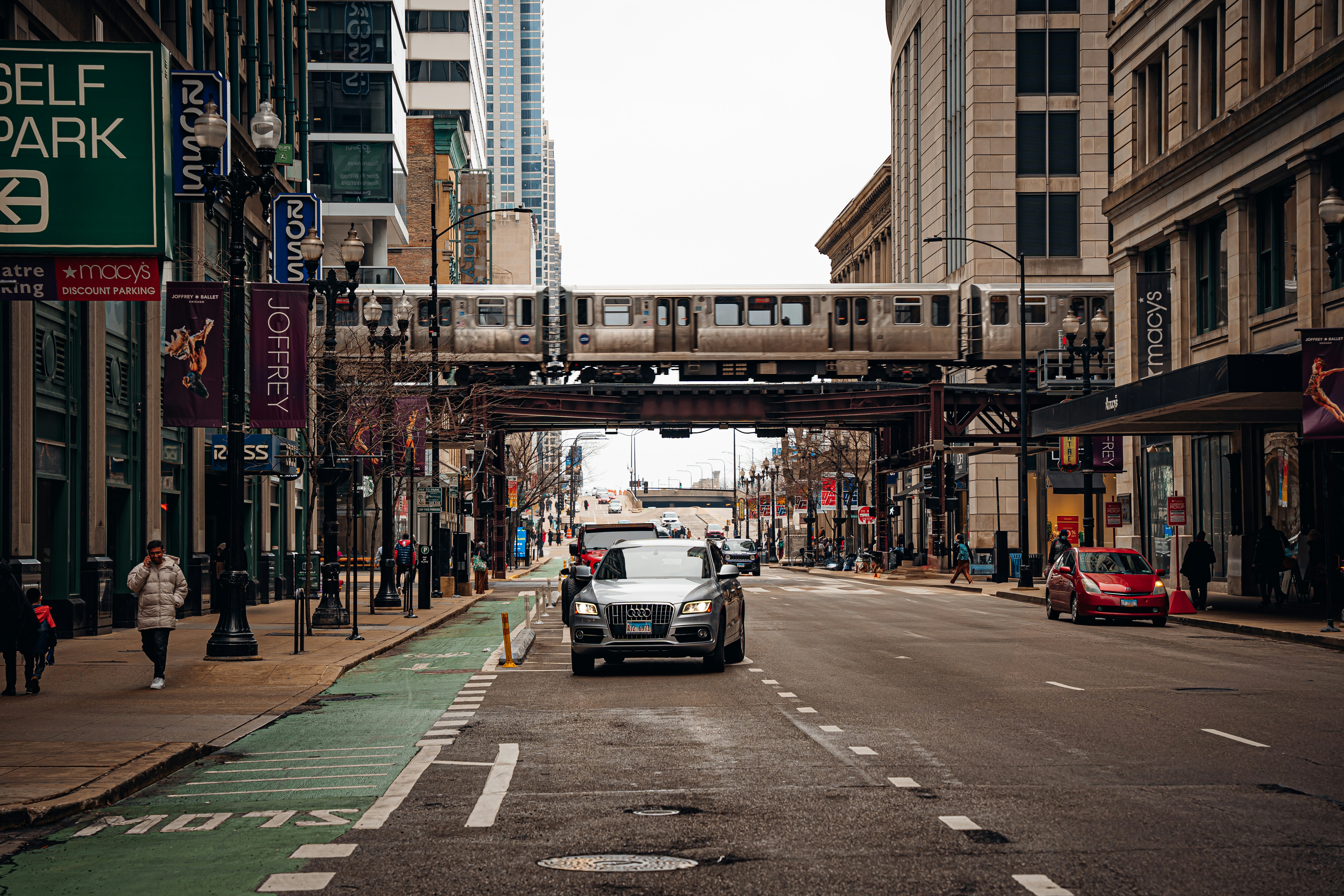 City street with cars below a passing elevated train and tall buildings lining the sidewalk.