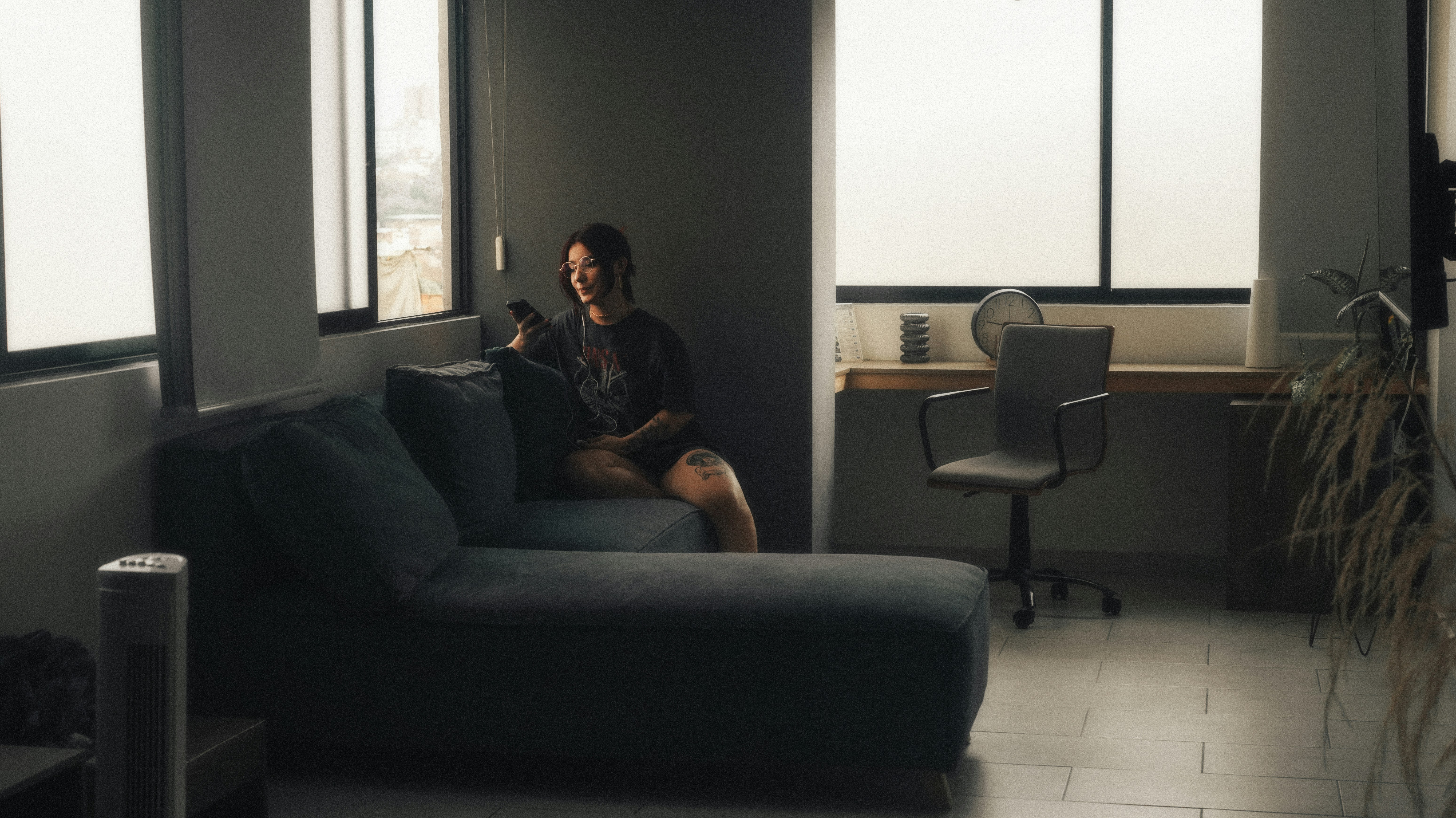 Person sitting on a couch in a softly lit room, reading a phone near large windows.