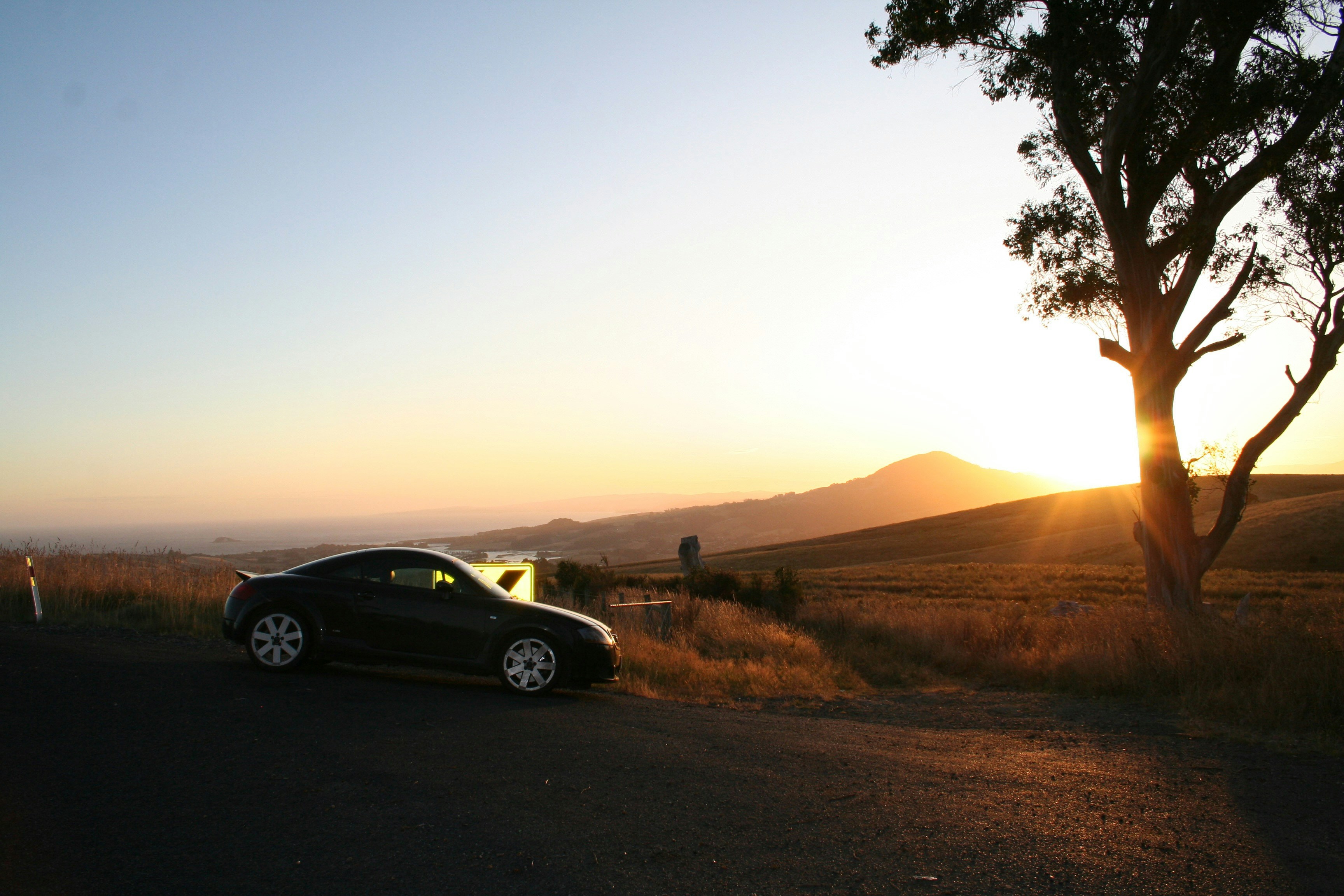 Car parked on a rural road at sunset with a distant mountain silhouette and a tree in the foreground.