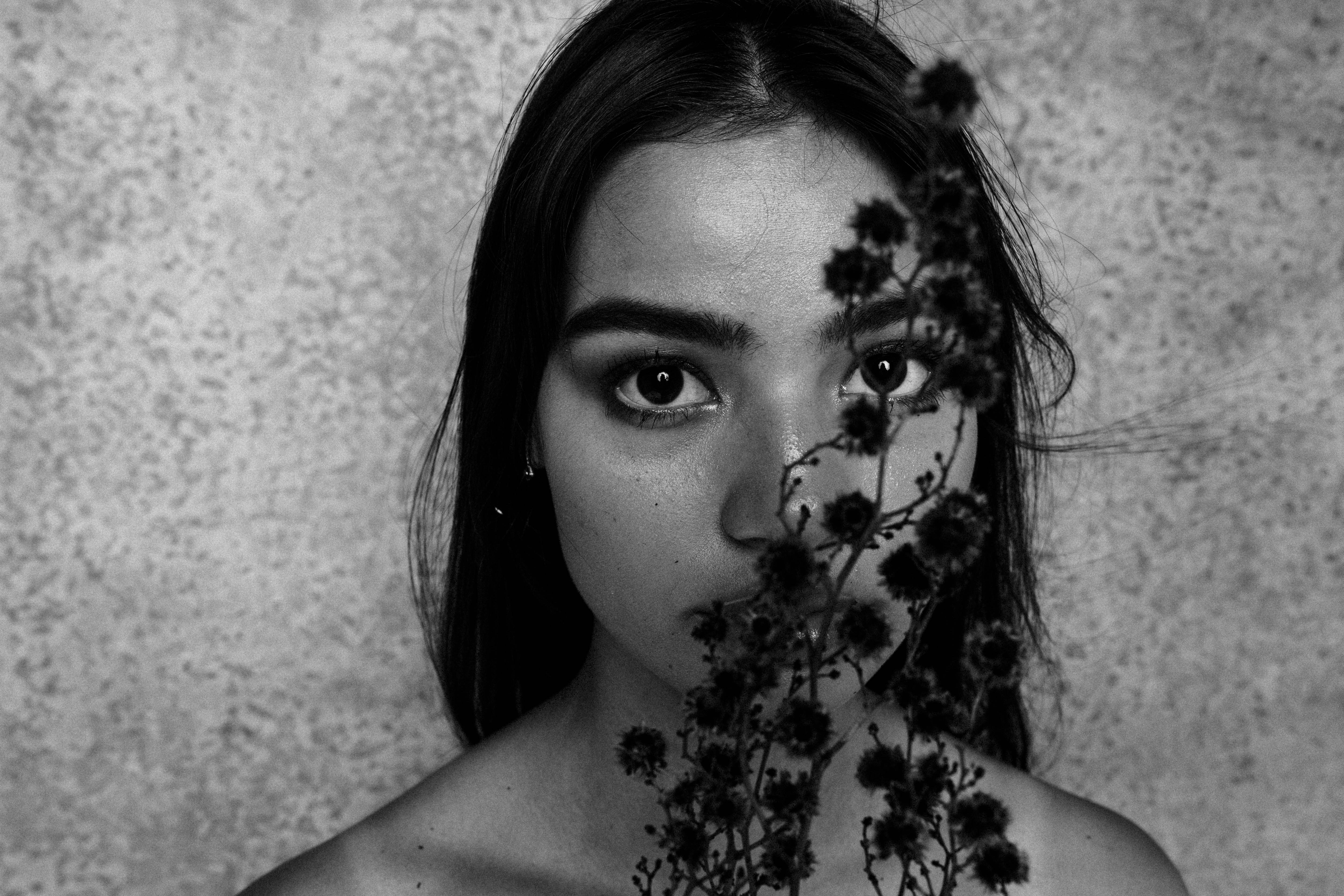 Monochrome portrait of a person with an intense gaze, partially obscured by dried flowers, set against a textured background.