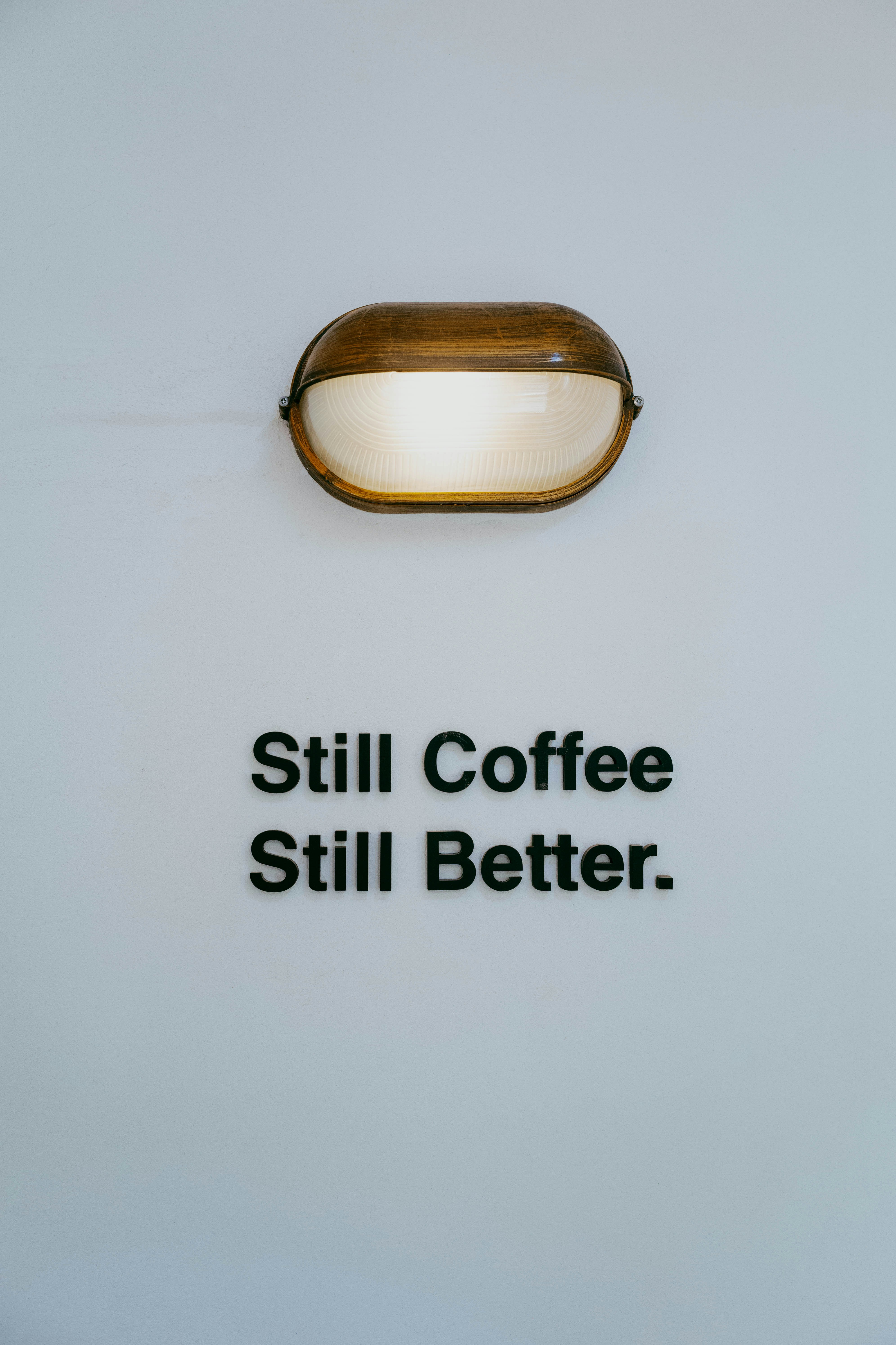 White wall with a wooden-framed light fixture above a bold, two-line slogan reading 'Still Coffee Still Better'.
