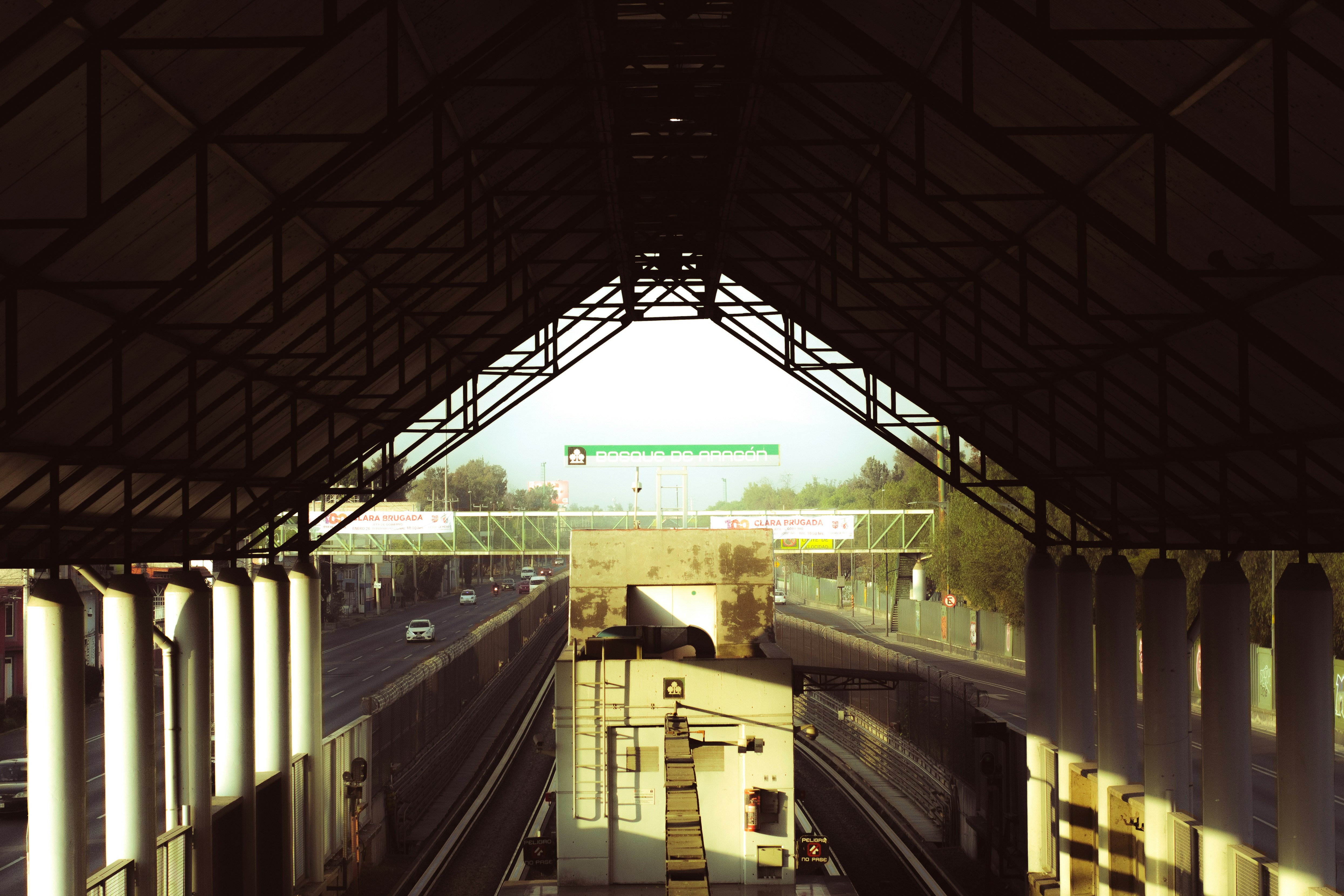 Sunlit train platform framed by geometric roof structure with road and trees in background.