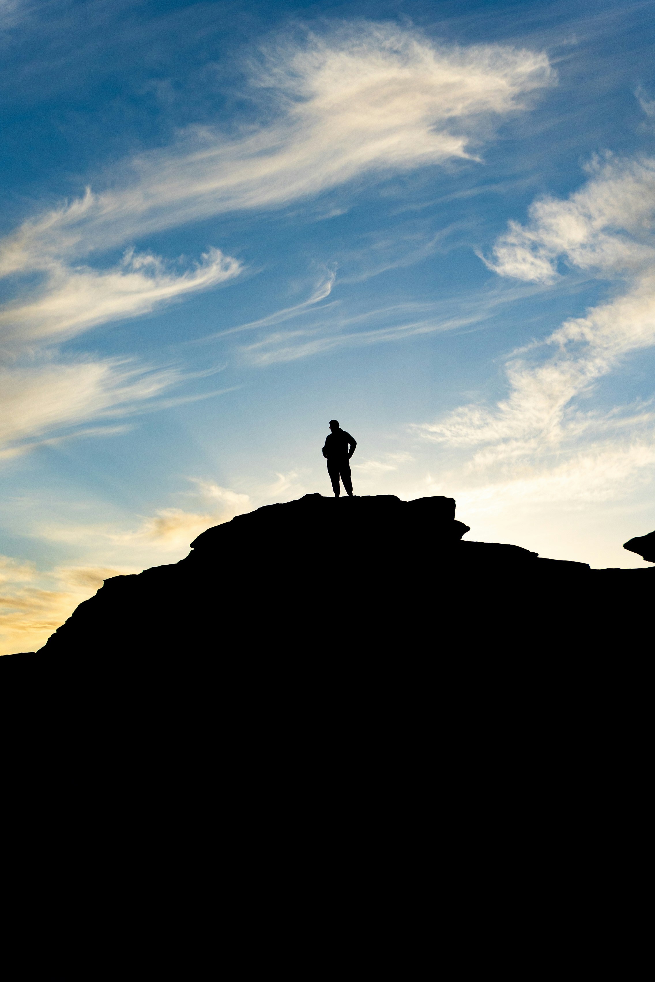 A lone figure stands atop a rocky outcrop, silhouetted against a vibrant sky filled with wispy clouds at dusk.