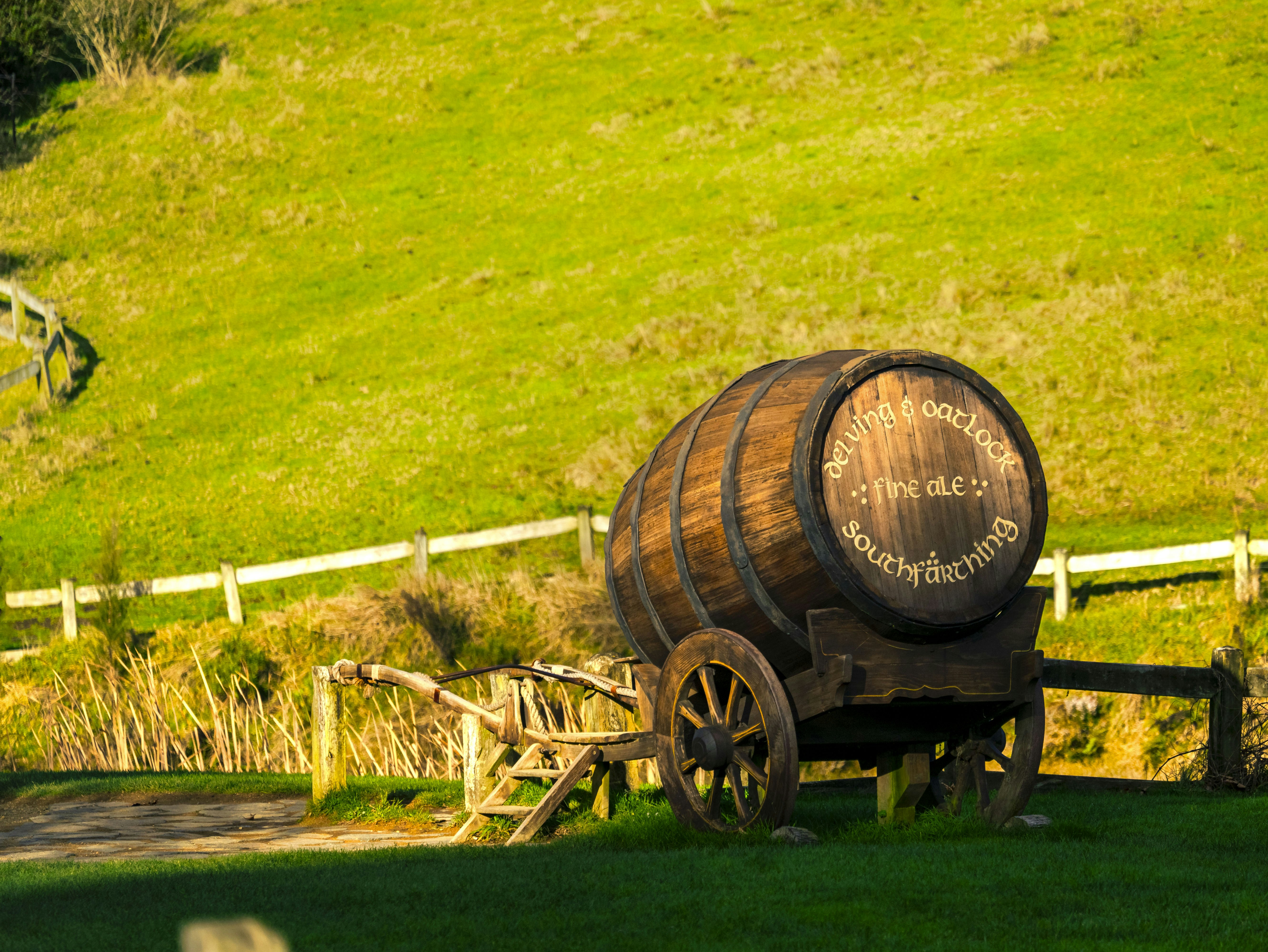 A weathered wooden ale barrel resting on a cart in a lush green meadow, surrounded by gentle hills and a wooden fence.