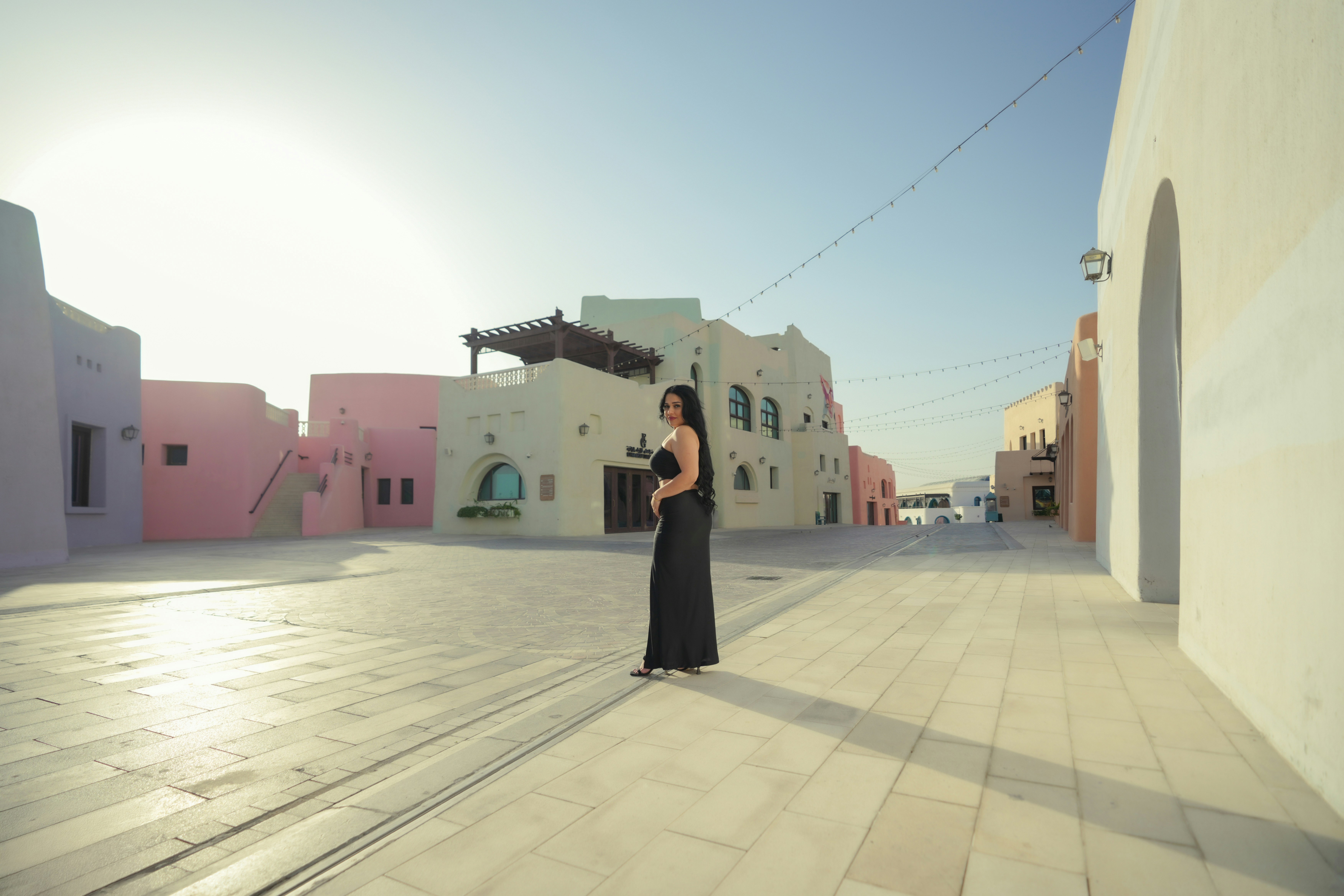 Person in a black dress stands in a sunlit plaza with pastel-colored buildings against a clear blue sky.