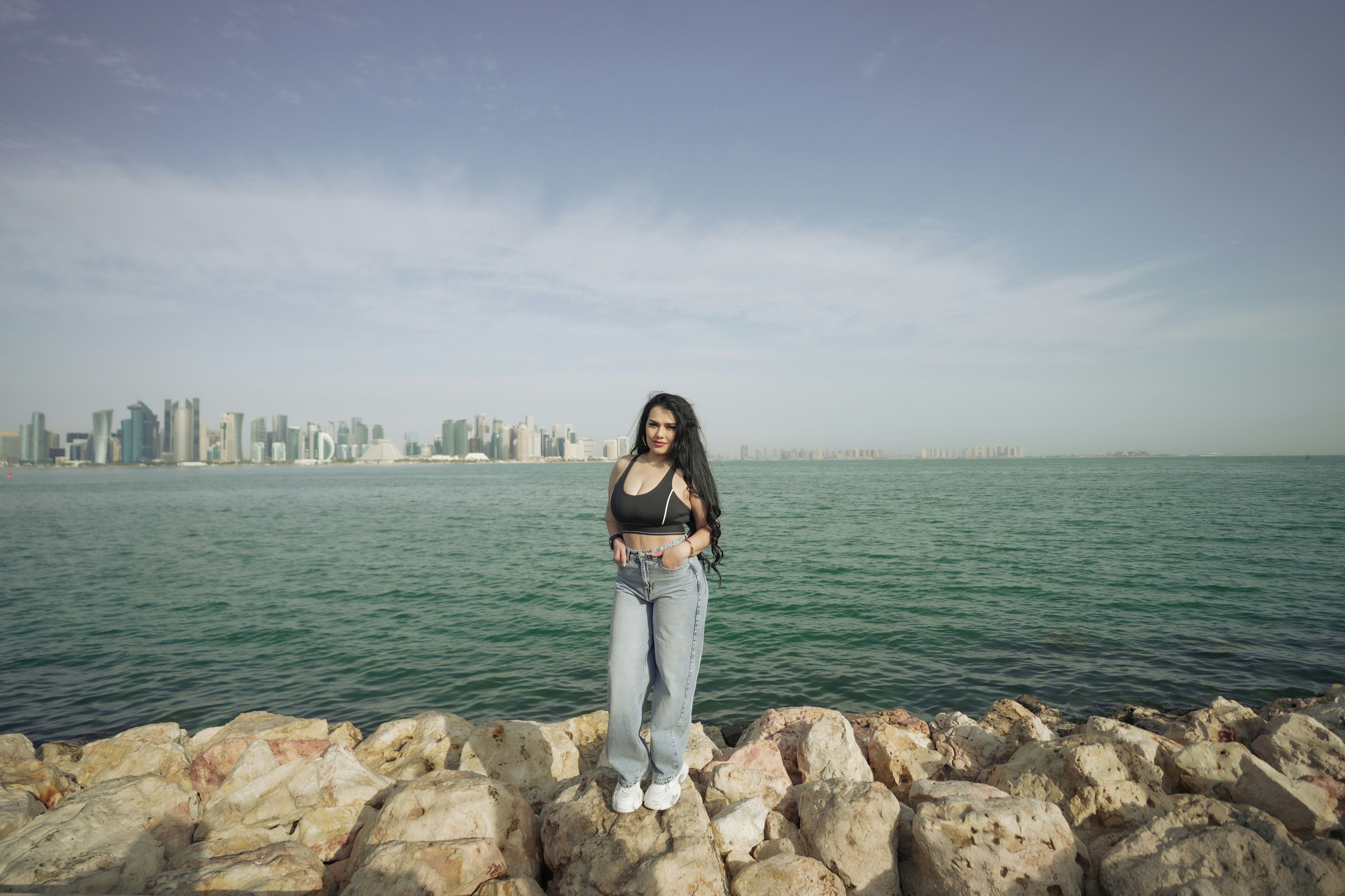 Woman standing on rocky shore with a modern city skyline in the background under a clear sky.