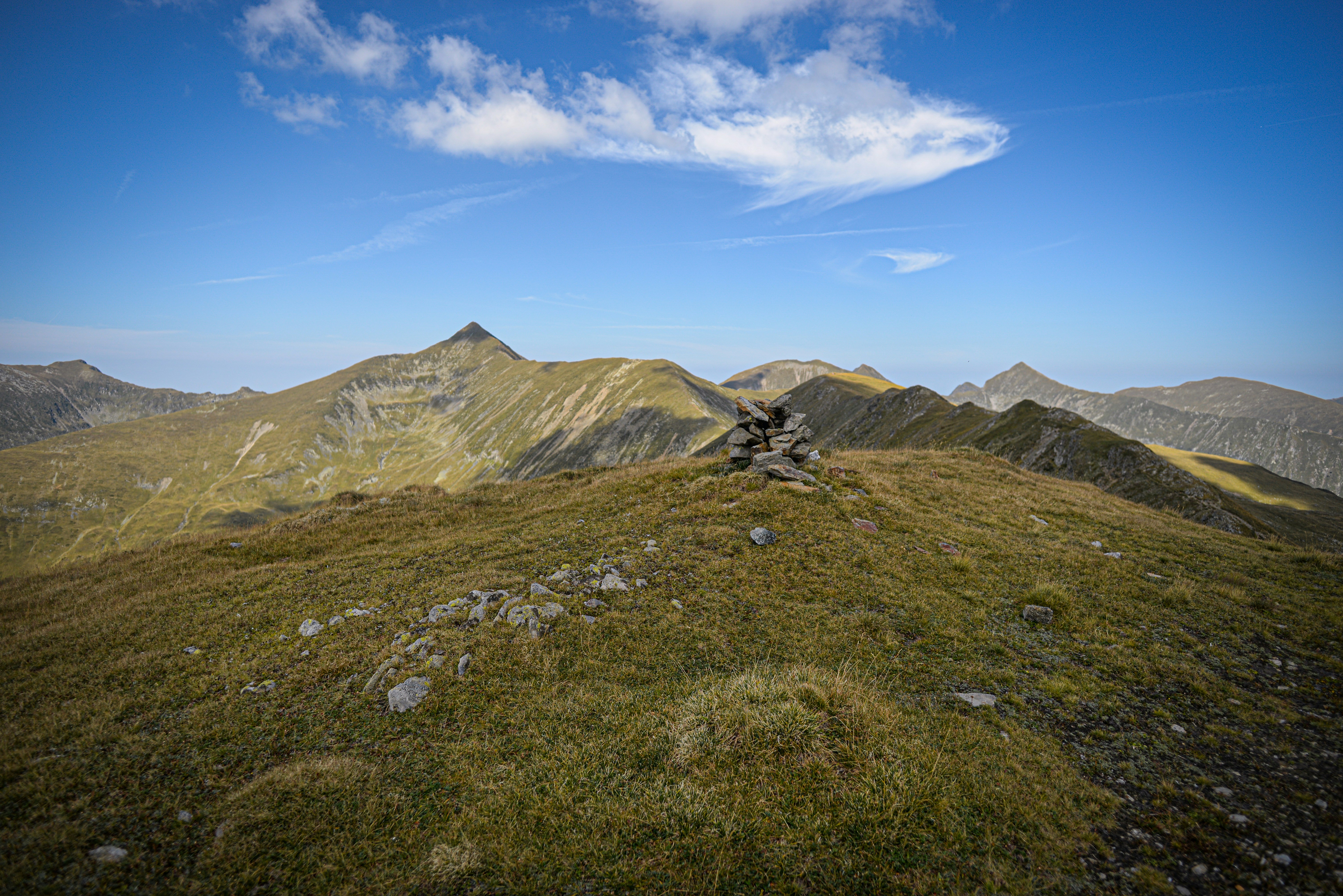 Mountain range beneath a vivid blue sky with a solitary cairn on a grassy ridge.