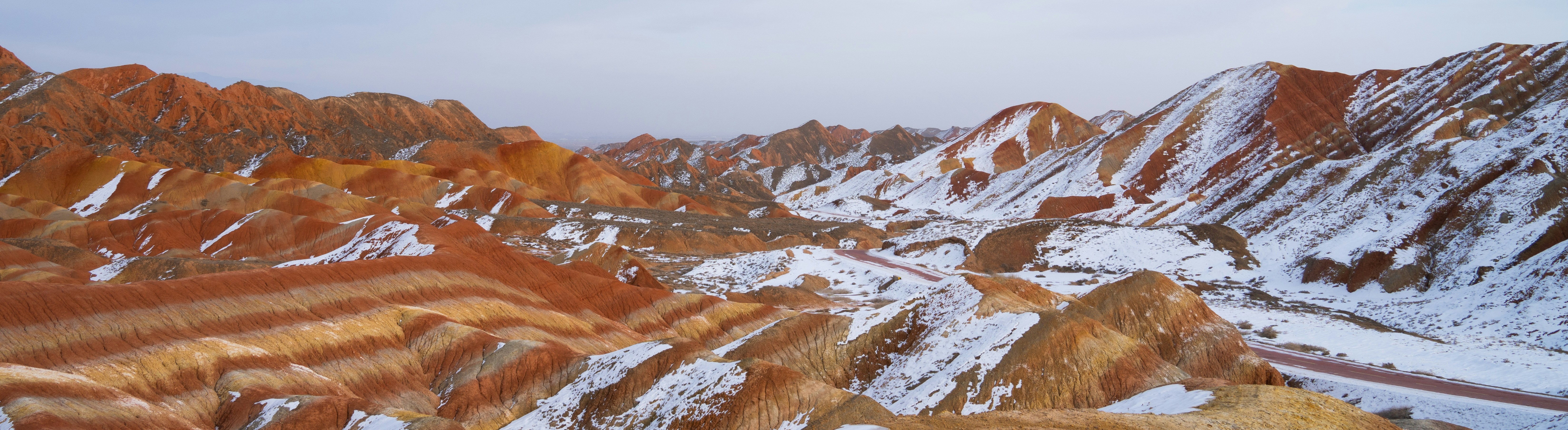 Multicolored hills with snow patches under a soft, overcast sky.