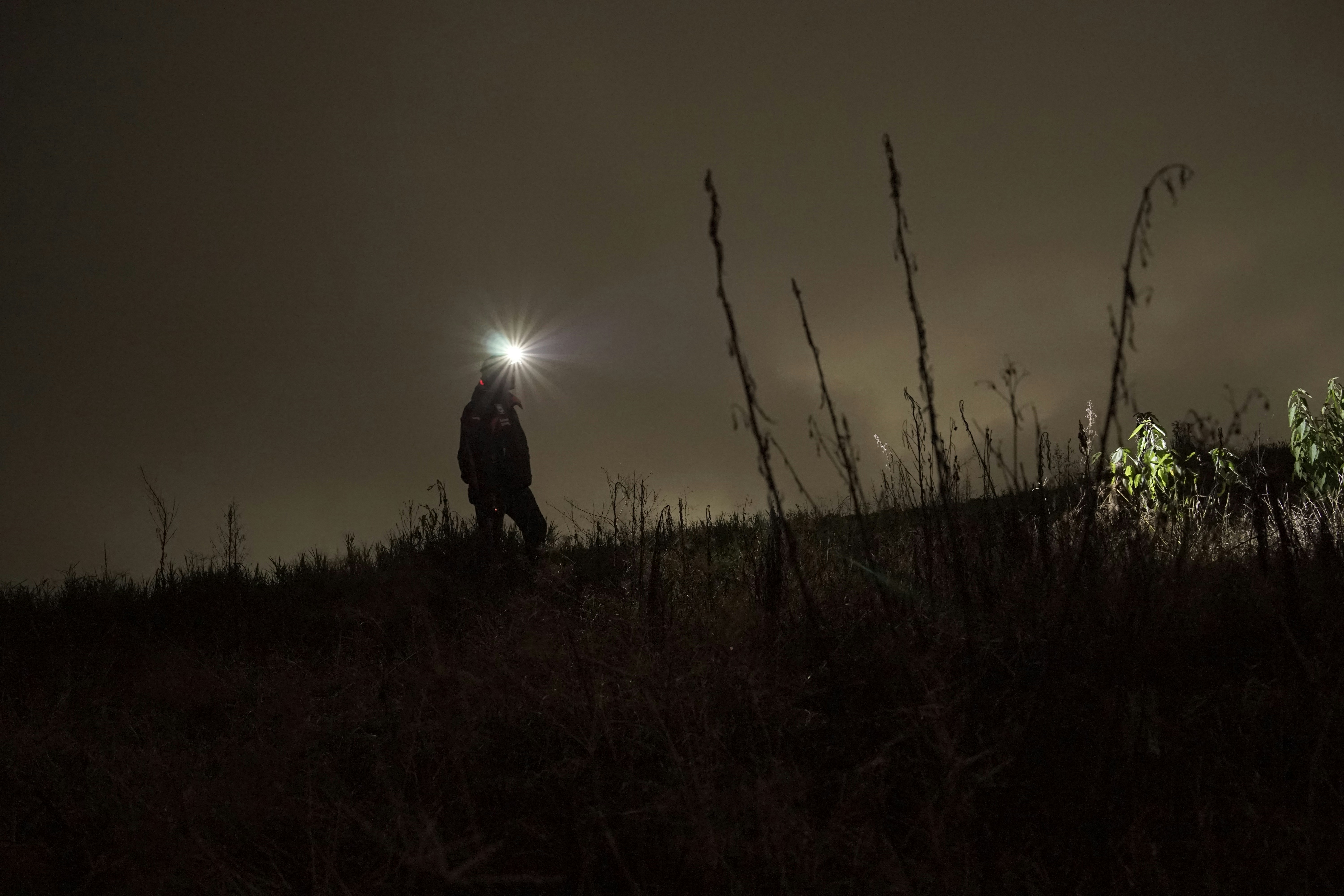 Silhouette of a person with a headlamp walking on a grassy hill at night.