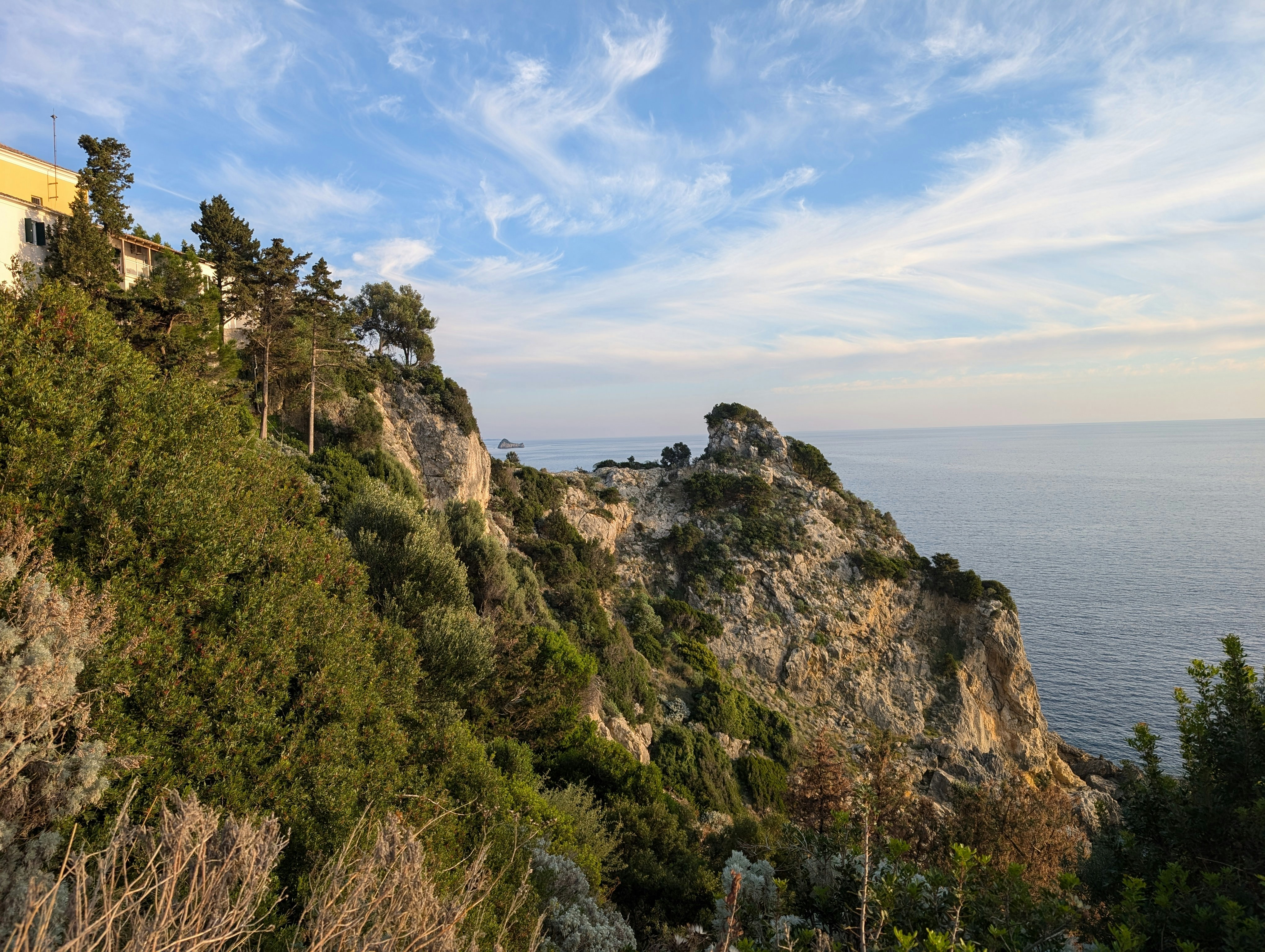Coastal cliffside landscape with rugged rocks, pine trees, and a building perched on the left. A calm sea stretches to the horizon beneath a clear sky during golden hour.