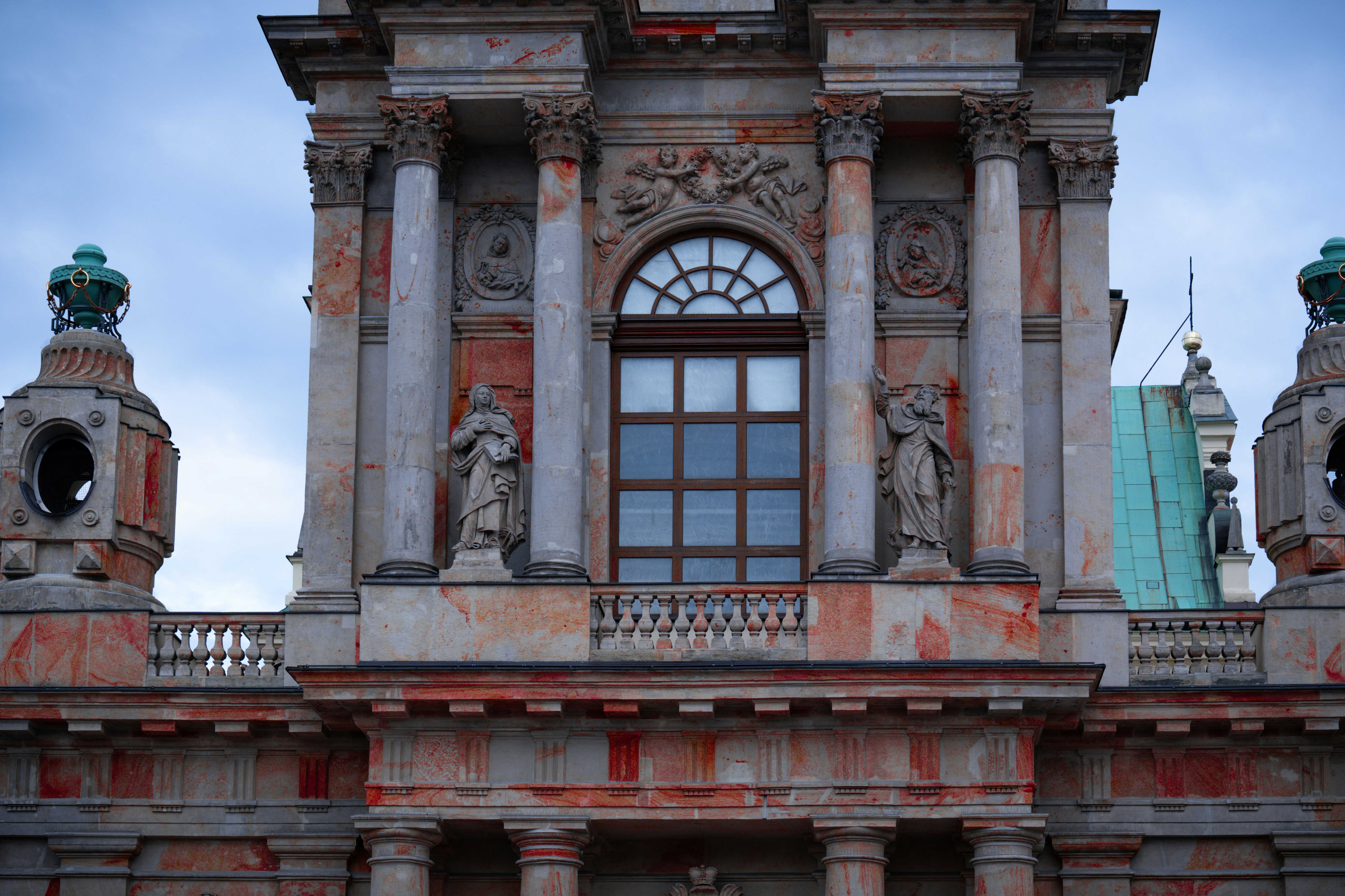 Ornate façade of a historic building with statues flanking a large arched window under a twilight sky.