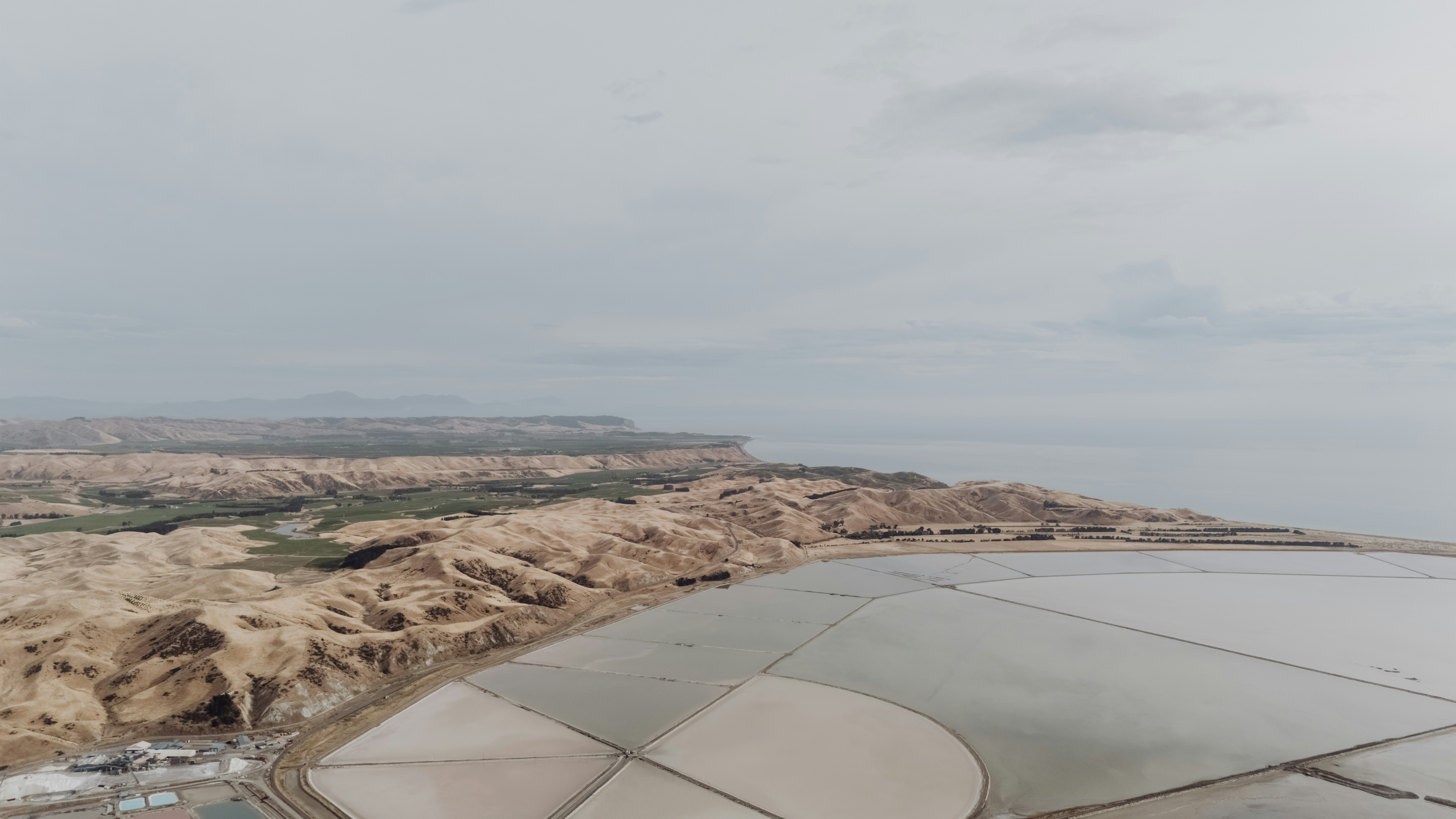 Expansive salt pans meet rolling hills under an overcast sky near the coast.