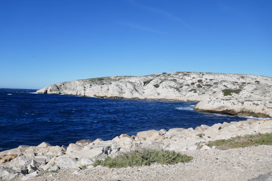 Bright rocky shoreline with white limestone cliffs curving around a deep blue bay under a clear sky. Foreground stones and scrub frame the coastal scene.