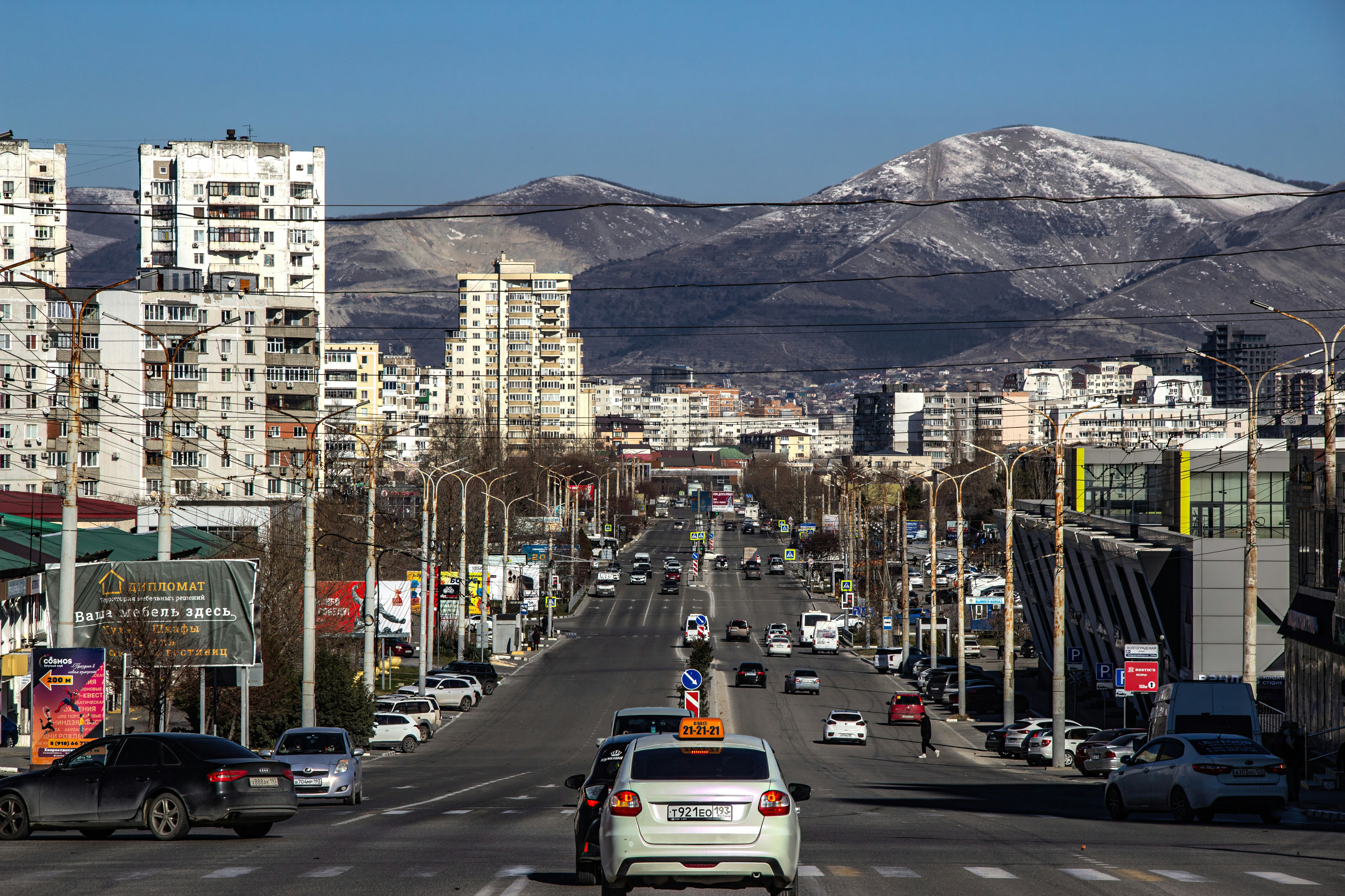 Bustling urban street lined with buildings leading to snow-capped mountains under a clear blue sky.