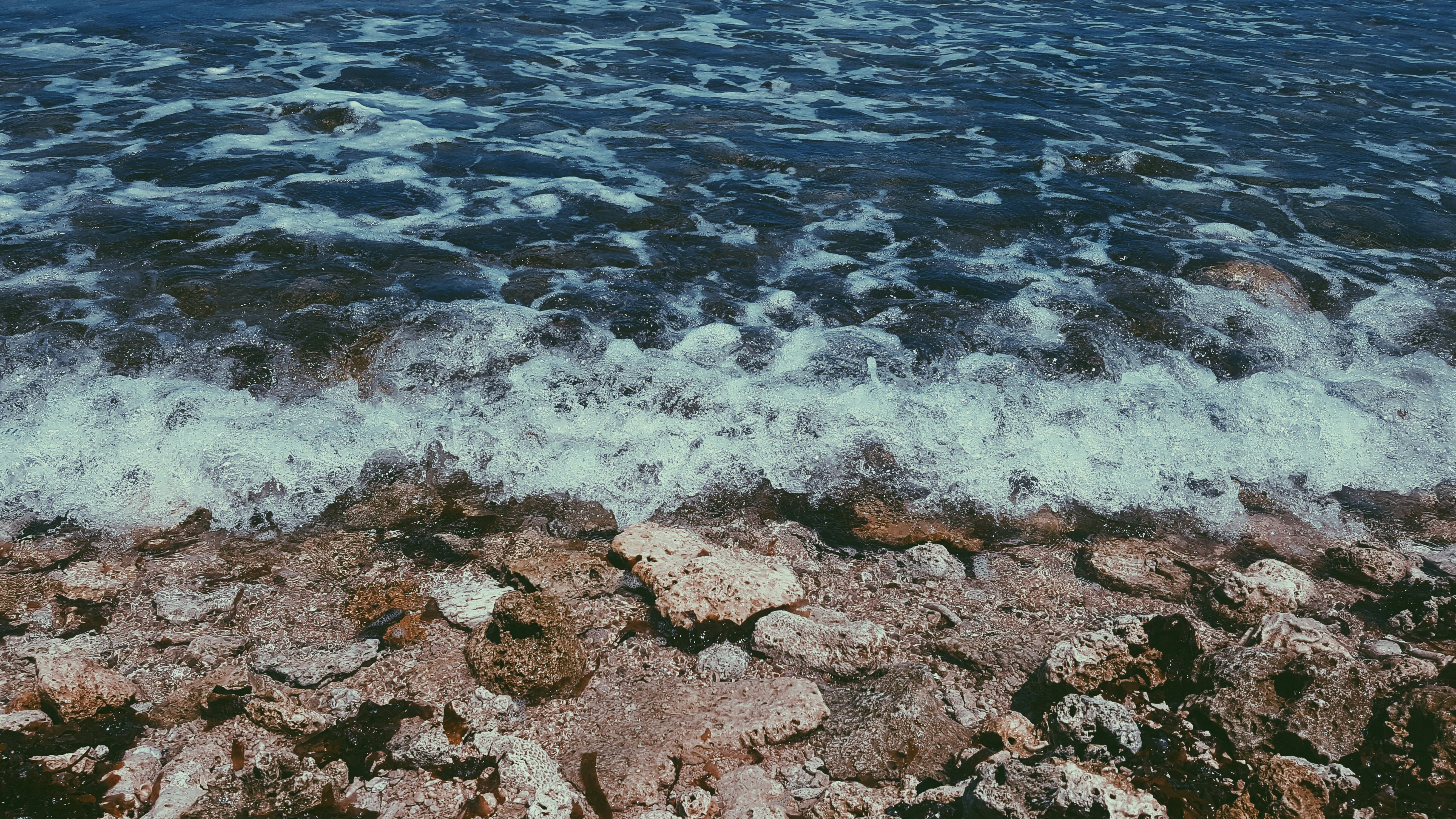 Waves gently wash over a rocky shoreline, contrasting the deep blue sea with textured earth tones under sunlight.