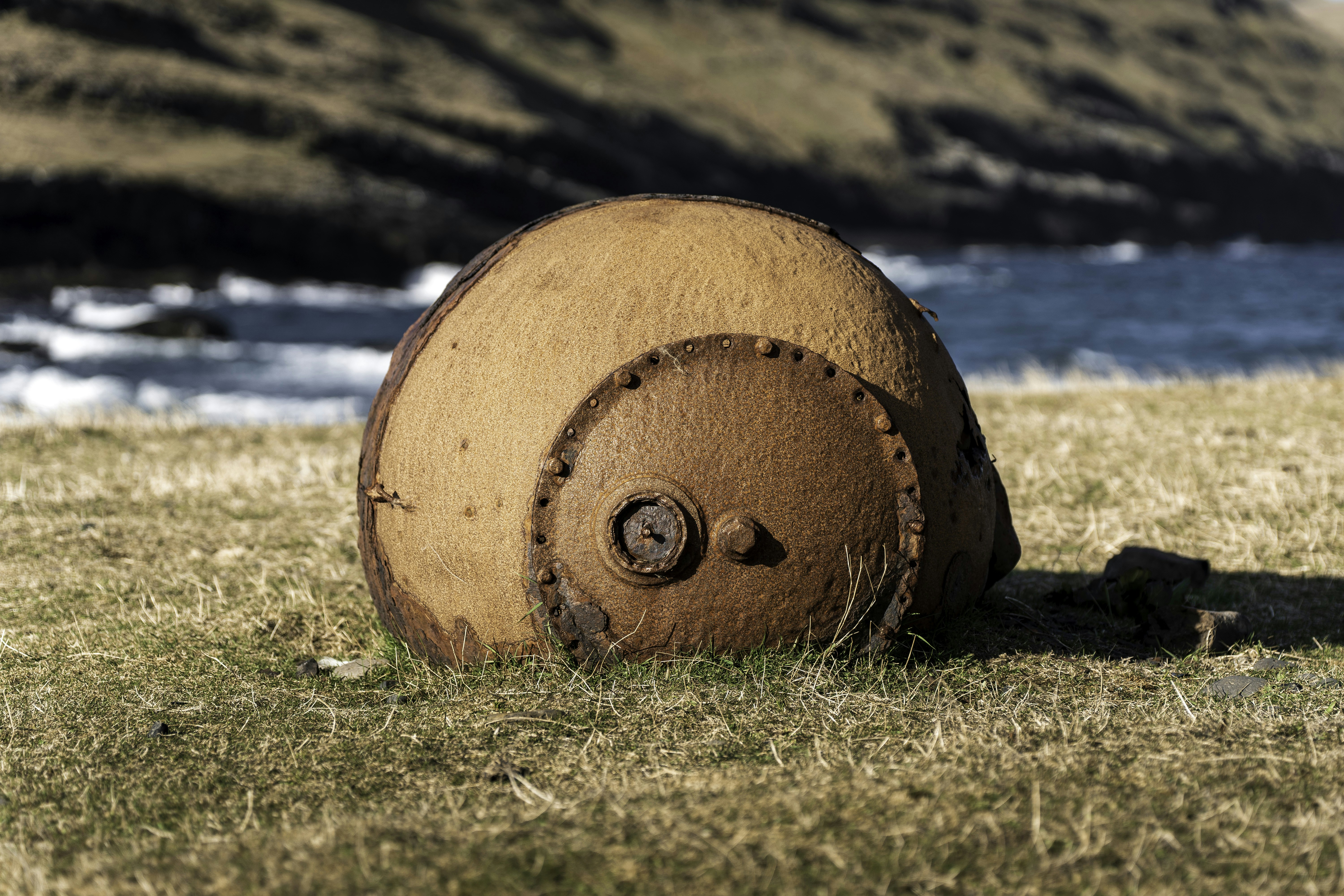 Weathered metal object resting on grassy terrain near the ocean, showcasing the passage of time and nature's elements.