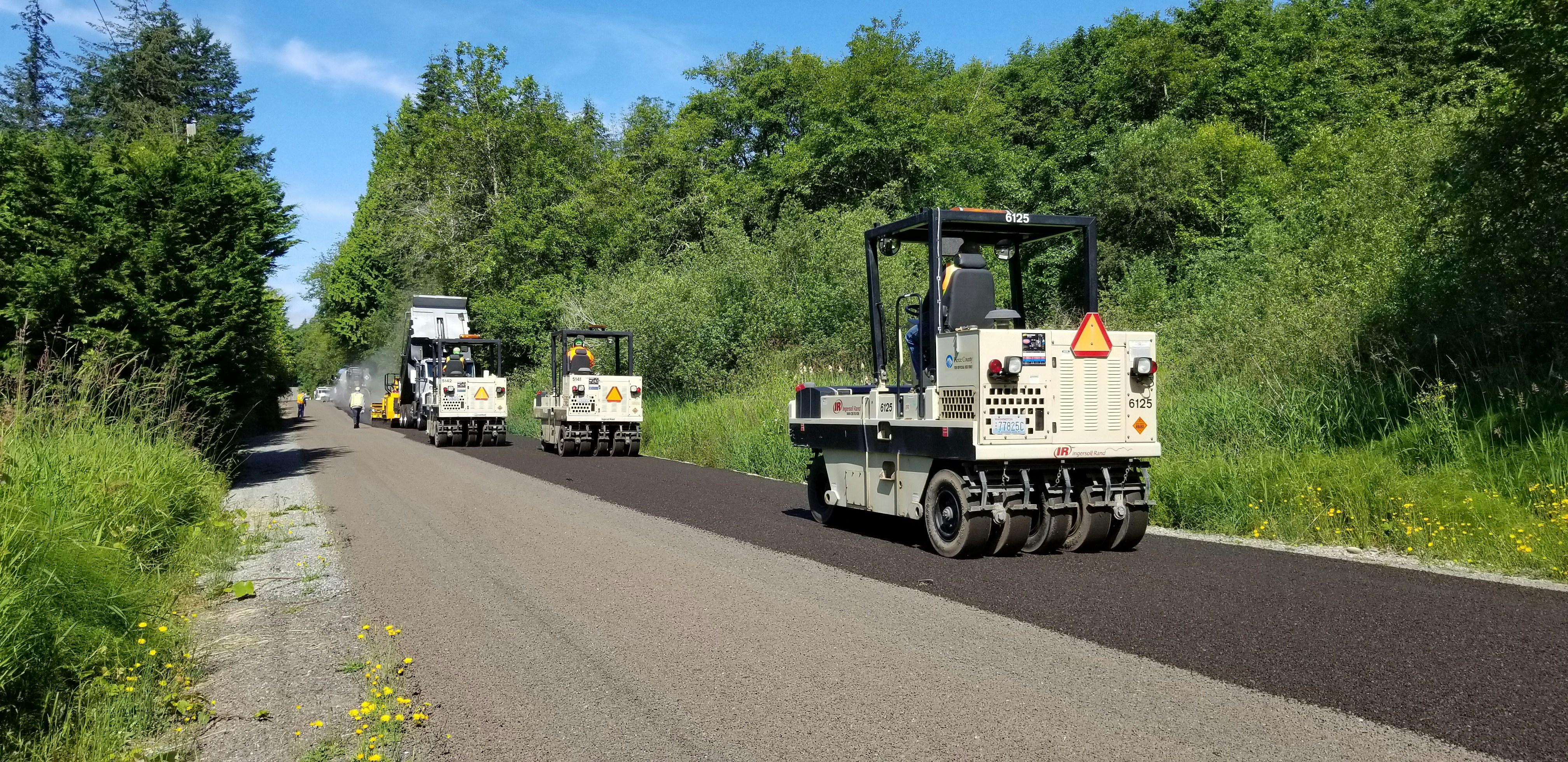 Paving machines lined up on a rural road surrounded by lush greenery under a clear blue sky.