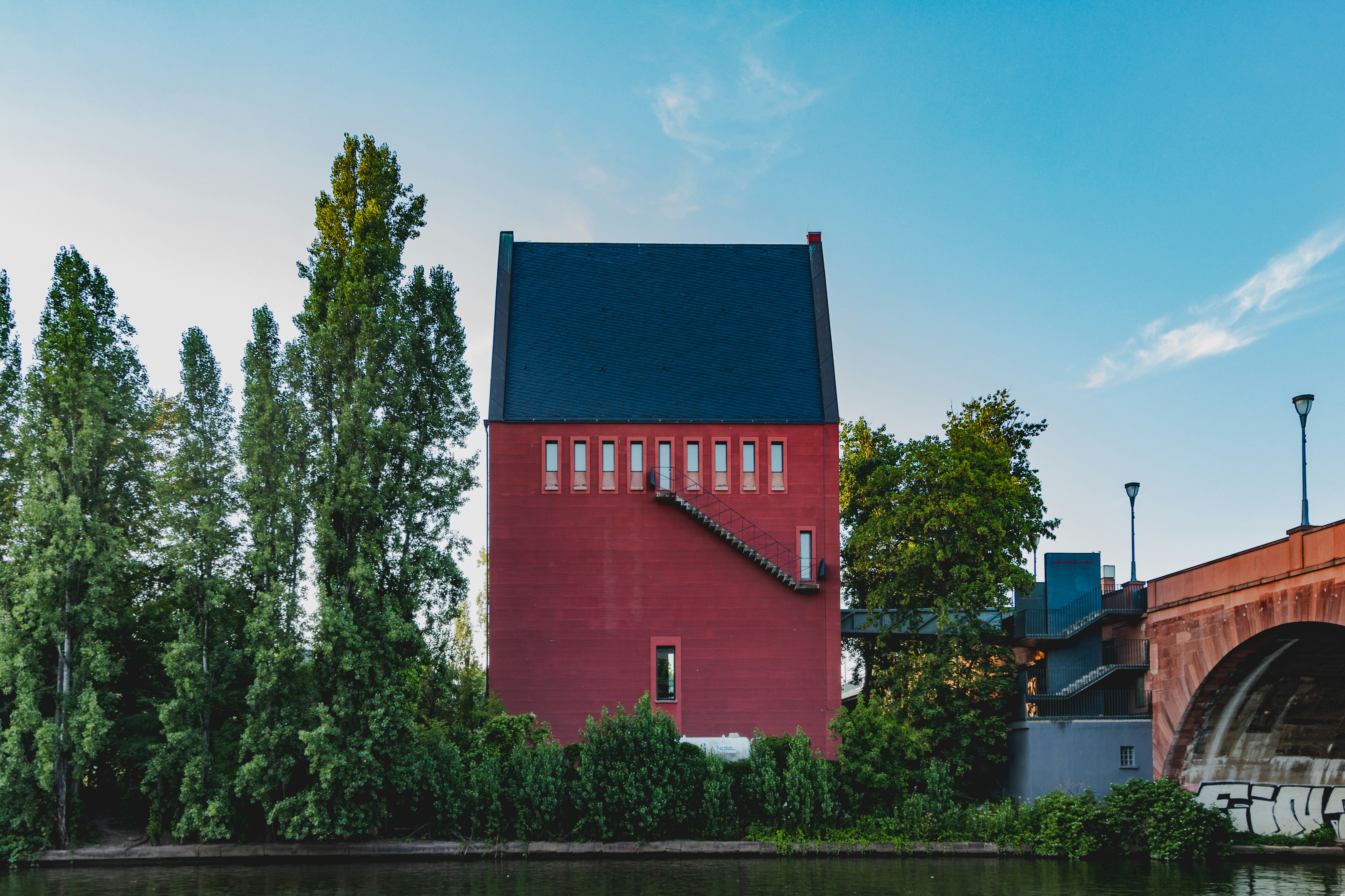 A striking red building with a unique sloped roof stands amidst lush greenery, reflecting modern architectural design. The structure is complemented by nearby trees and a bridge.