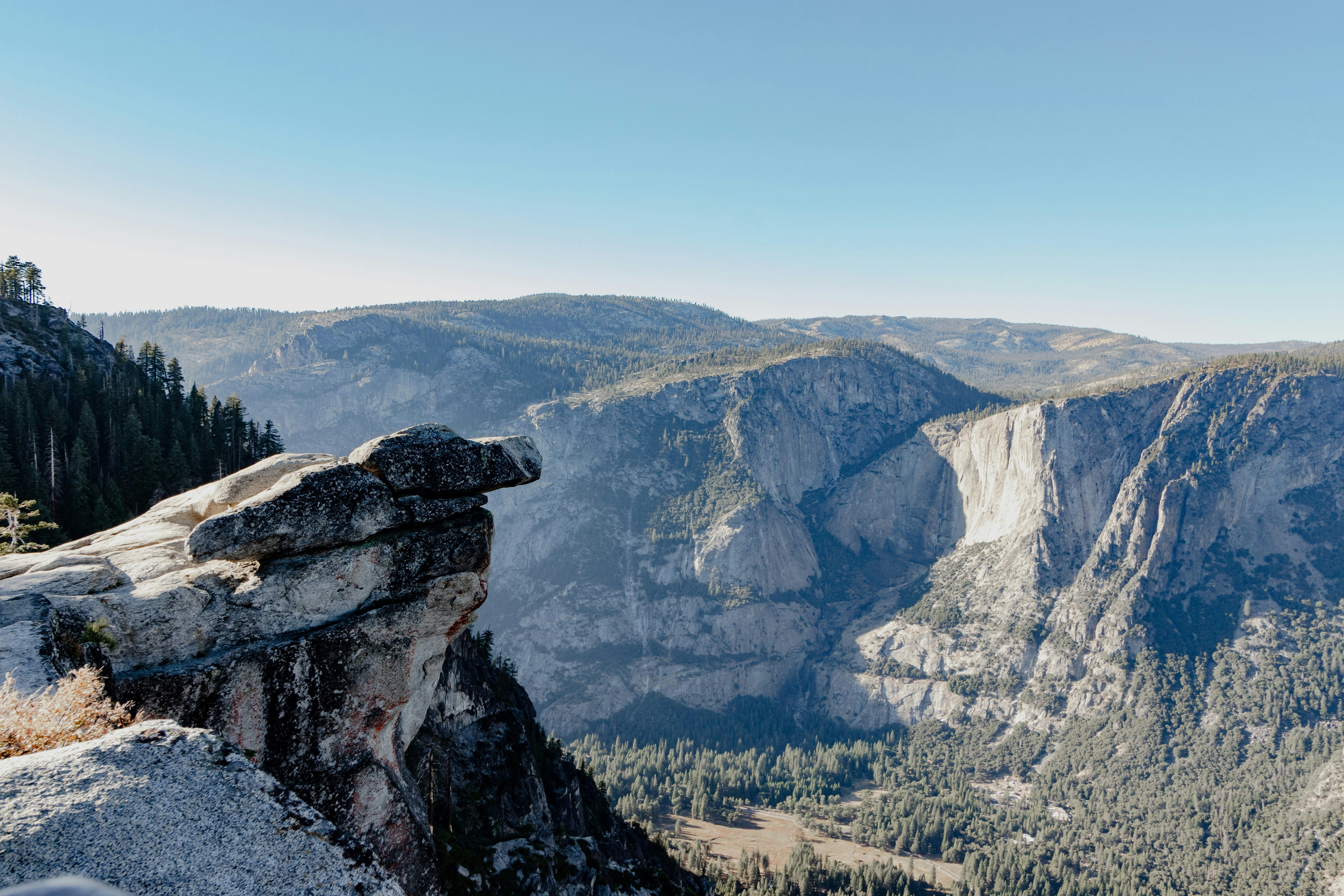 Dramatic rock ledge extends over a sunlit canyon, with layered cliffs and dense forests stretching into the distance under a clear blue sky.