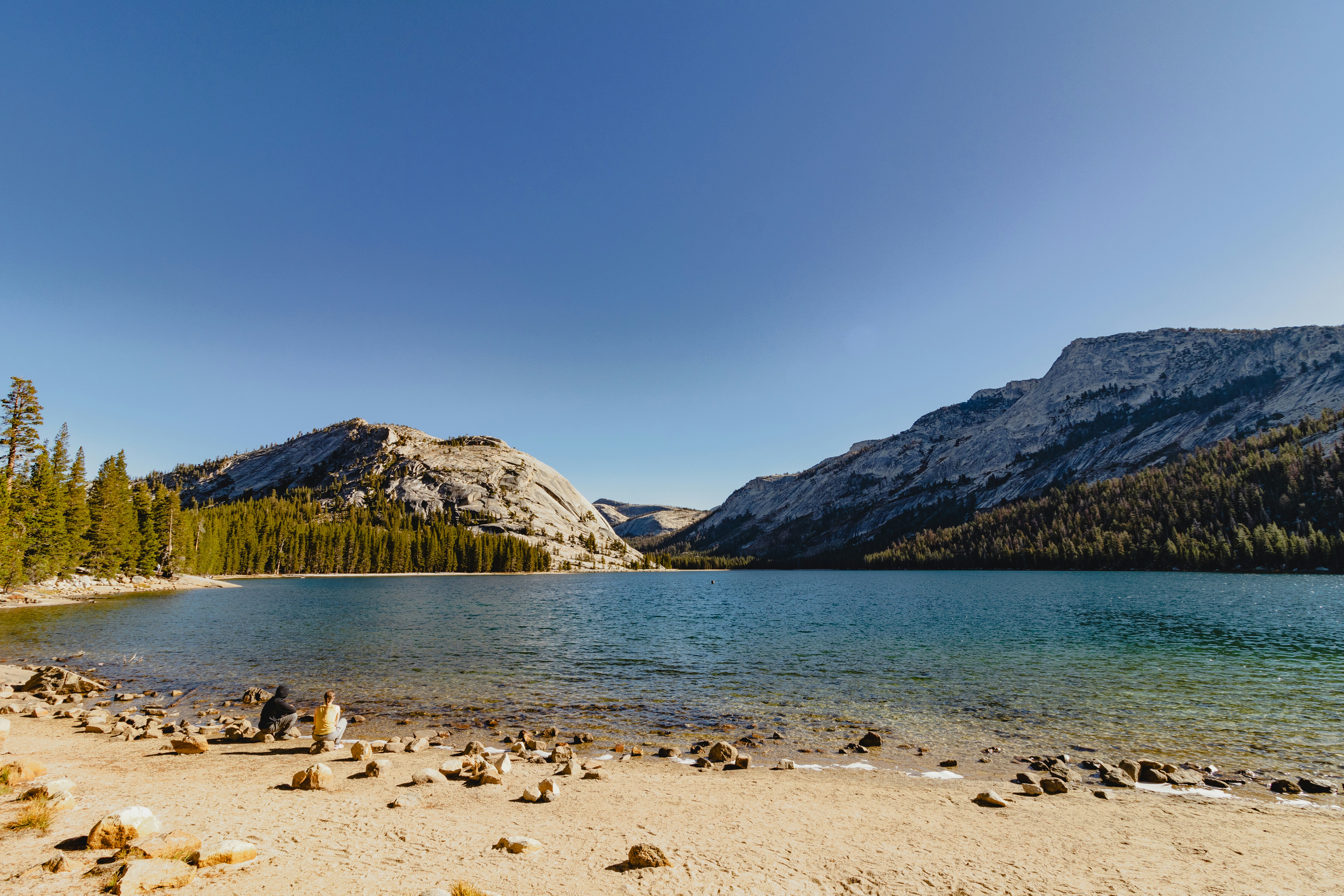 Sandy lakeshore with scattered rocks leading to a clear blue lake, flanked by forested mountains under a bright sky.