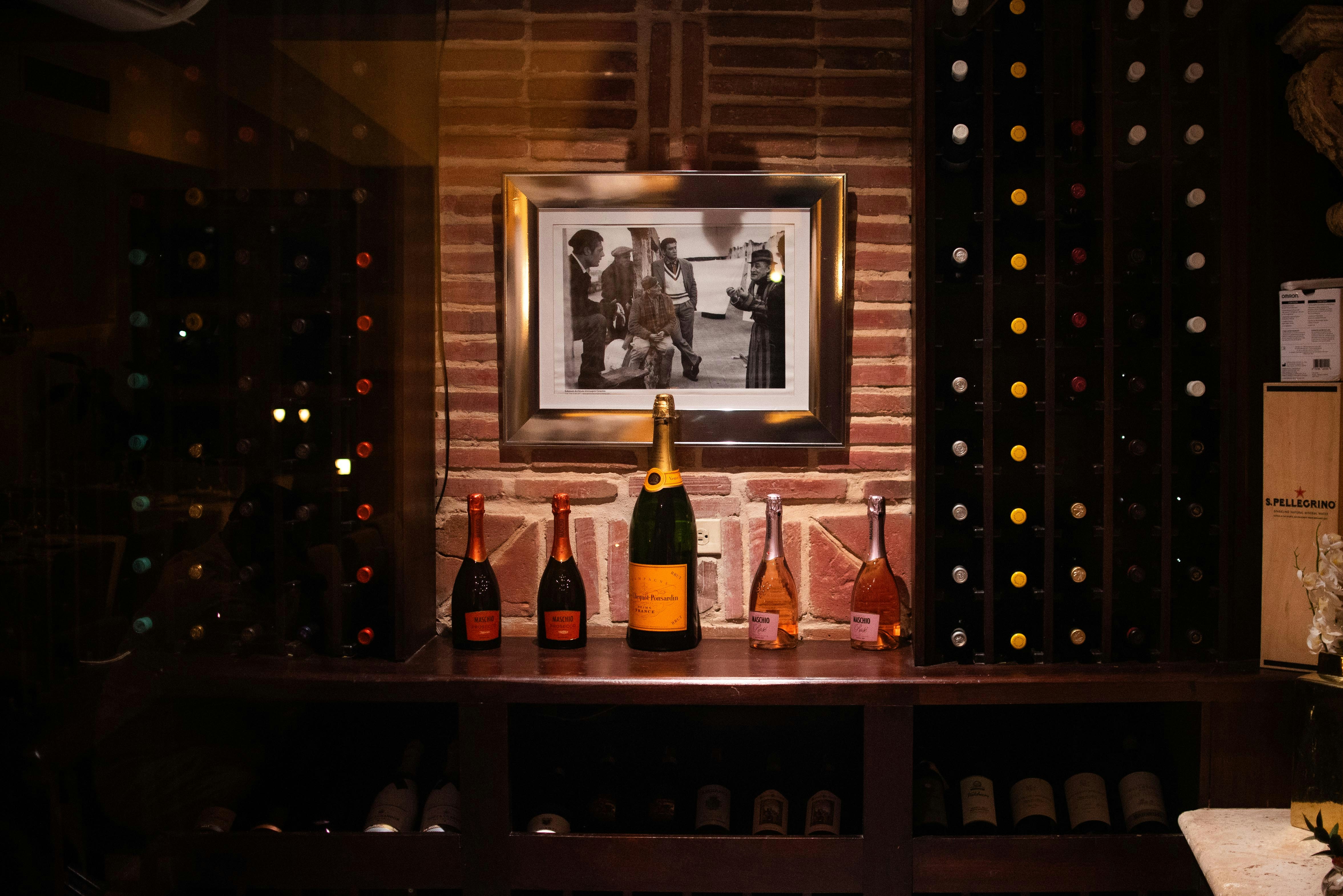 Dimly lit wine cellar with an array of bottles on a dark wooden table against a brick wall, featuring a framed black-and-white photograph.