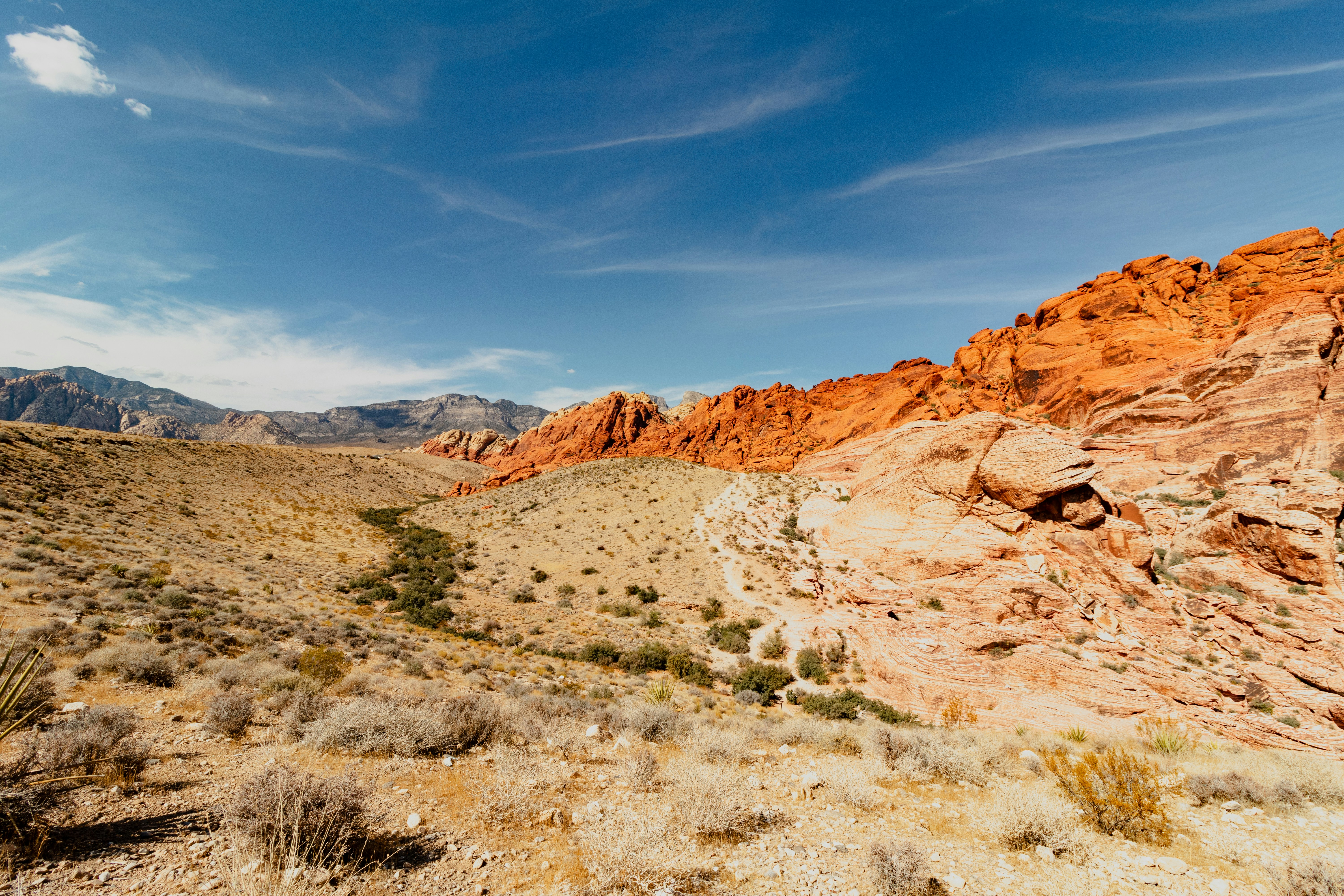 Vibrant red rock formations rise against a clear blue sky in a desert landscape, with earthy tones and scattered vegetation.