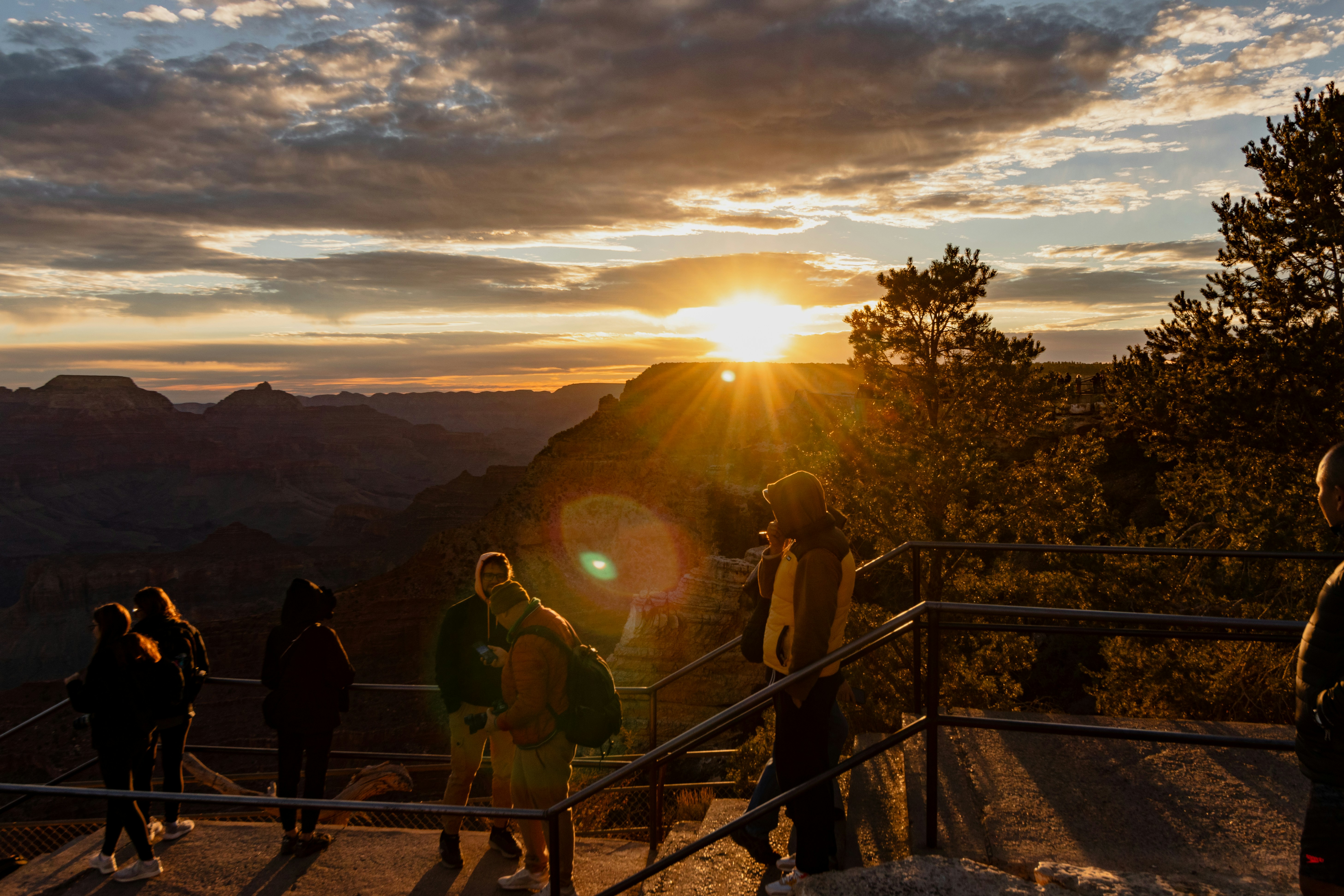 People silhouetted against a golden sunset at a canyon overlook, with dramatic clouds and rugged landscape.