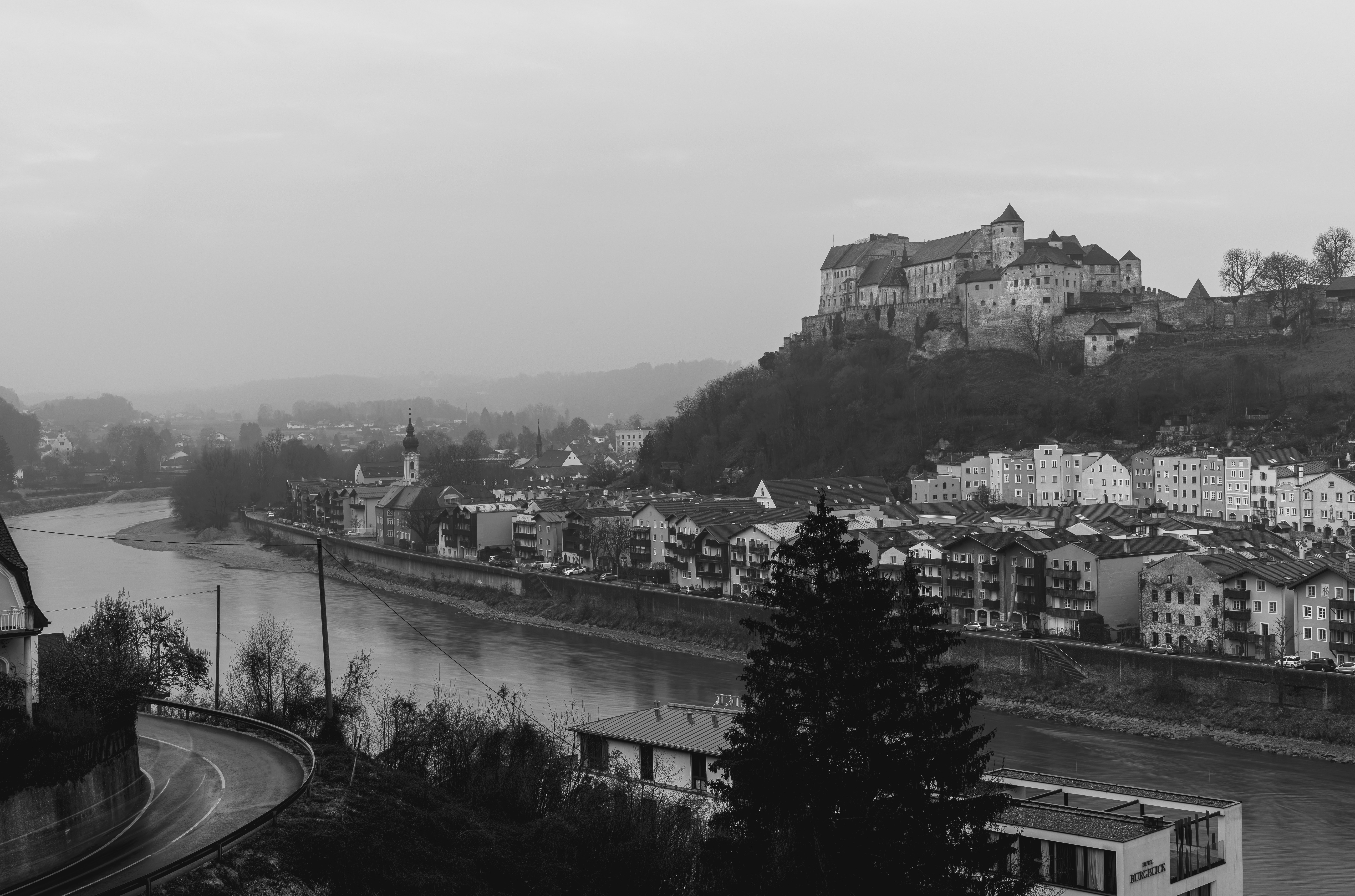 Historic castle on a hill overlooking a town and winding river under a misty sky.