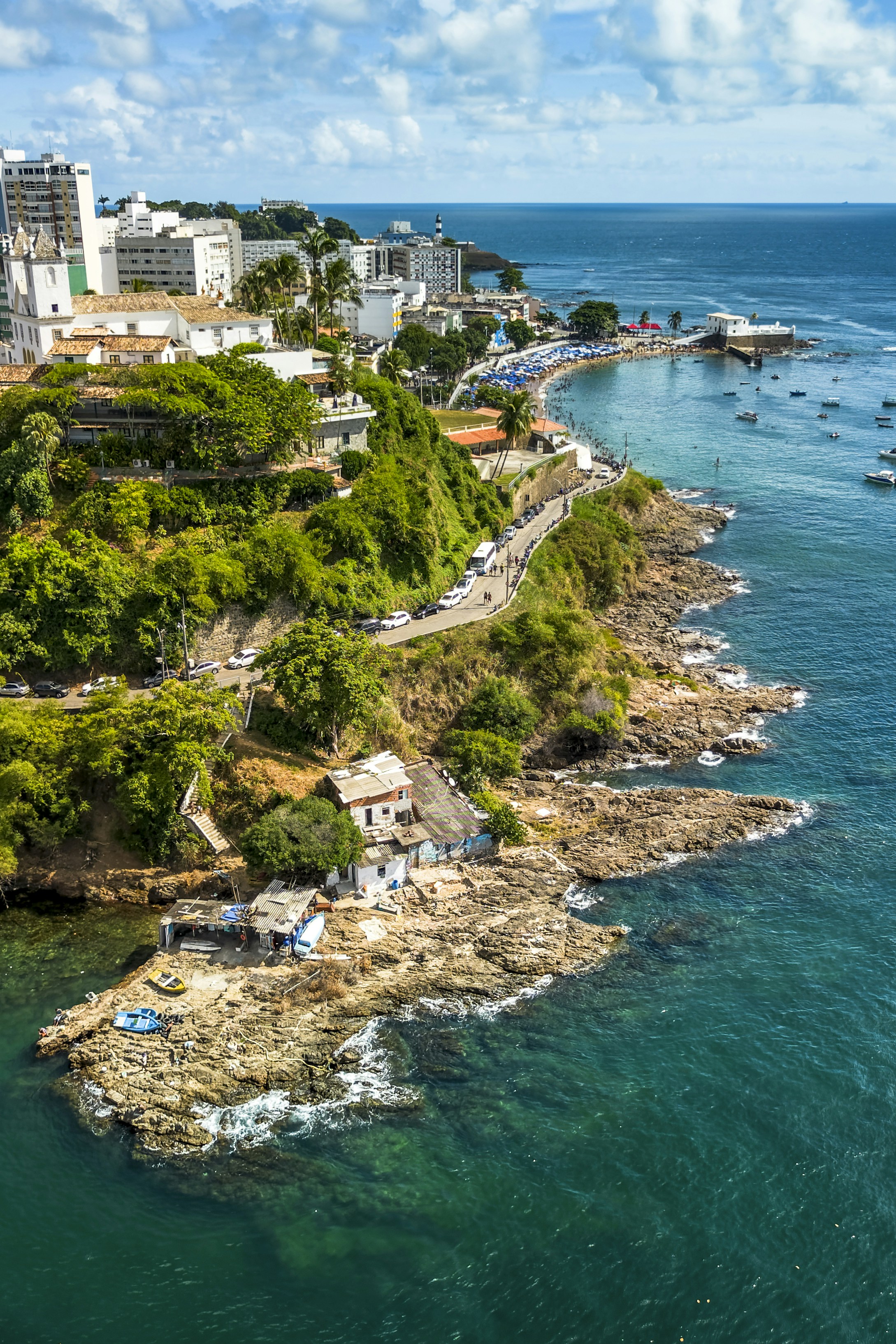 Aerial view of a vibrant coastline with lush greenery and urban buildings meeting the deep blue ocean under a partly cloudy sky.