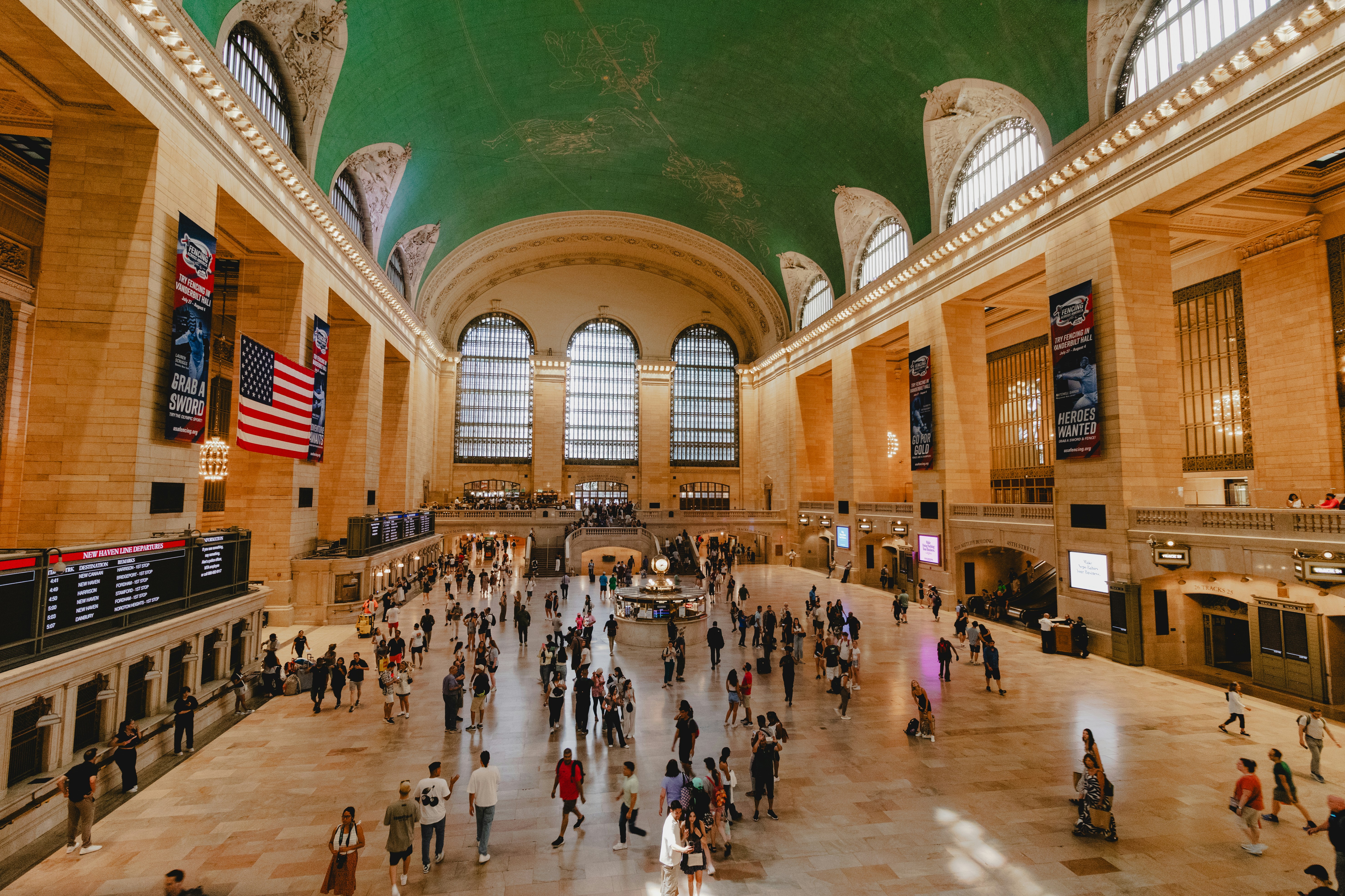 Crowded terminal hall with intricate green ceiling and bright natural light pouring through arched windows.