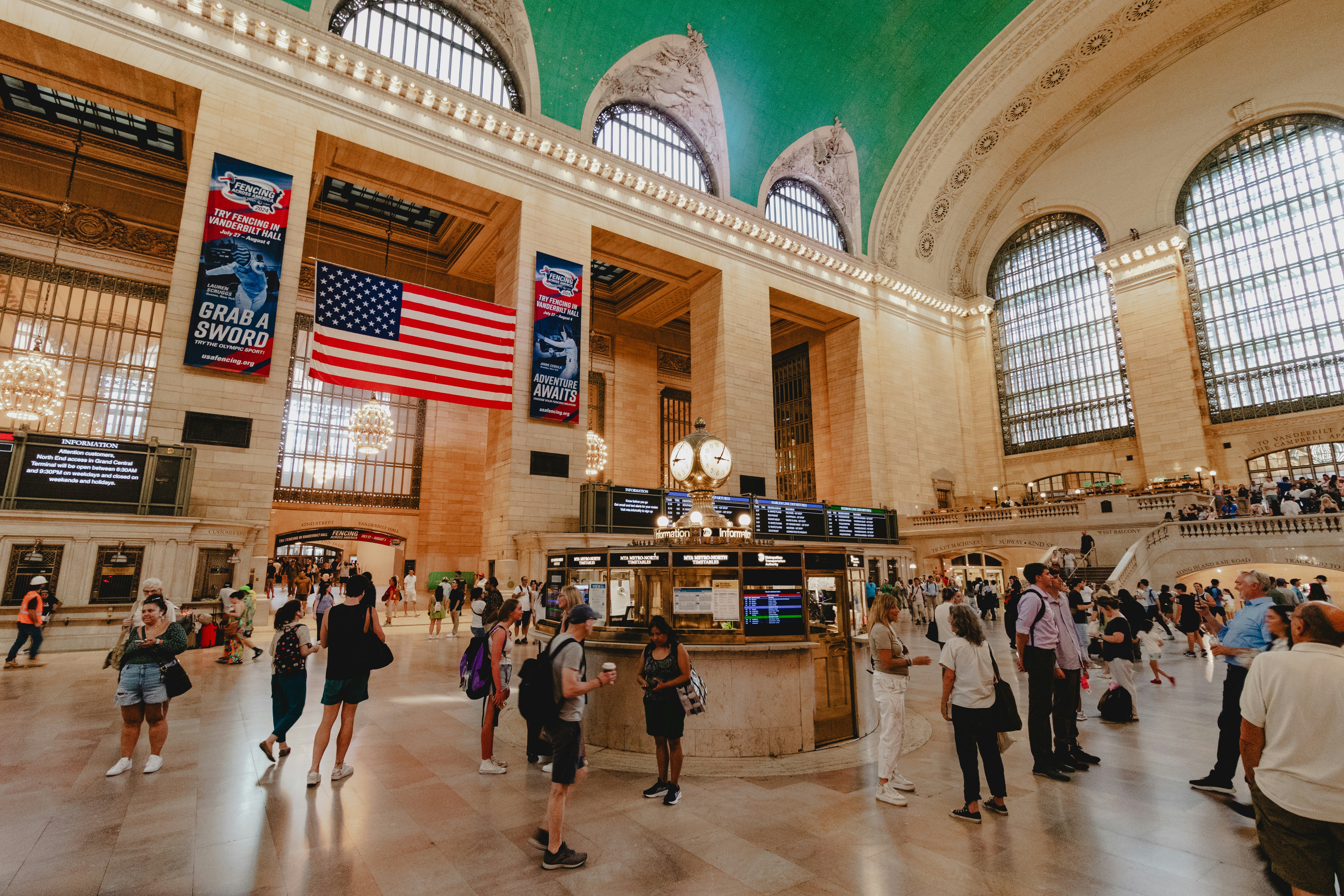 Crowd gathers under the iconic American flag and arched windows of Grand Central Terminal, with warm stone tones contrasting the cool green ceiling.