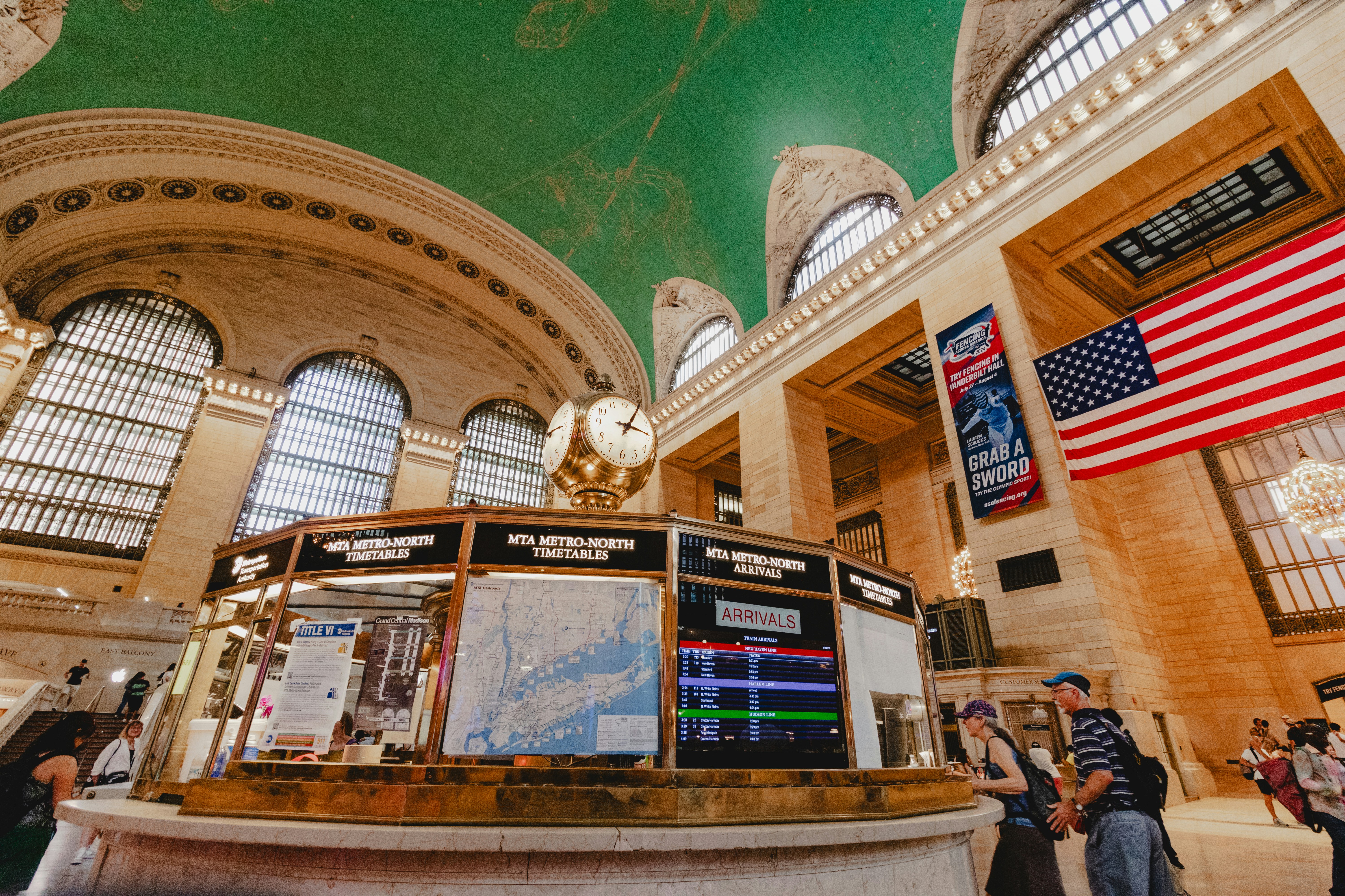 Grand Central Terminal's central information booth with a golden clock, beneath a vivid green ceiling with celestial murals and arched windows.