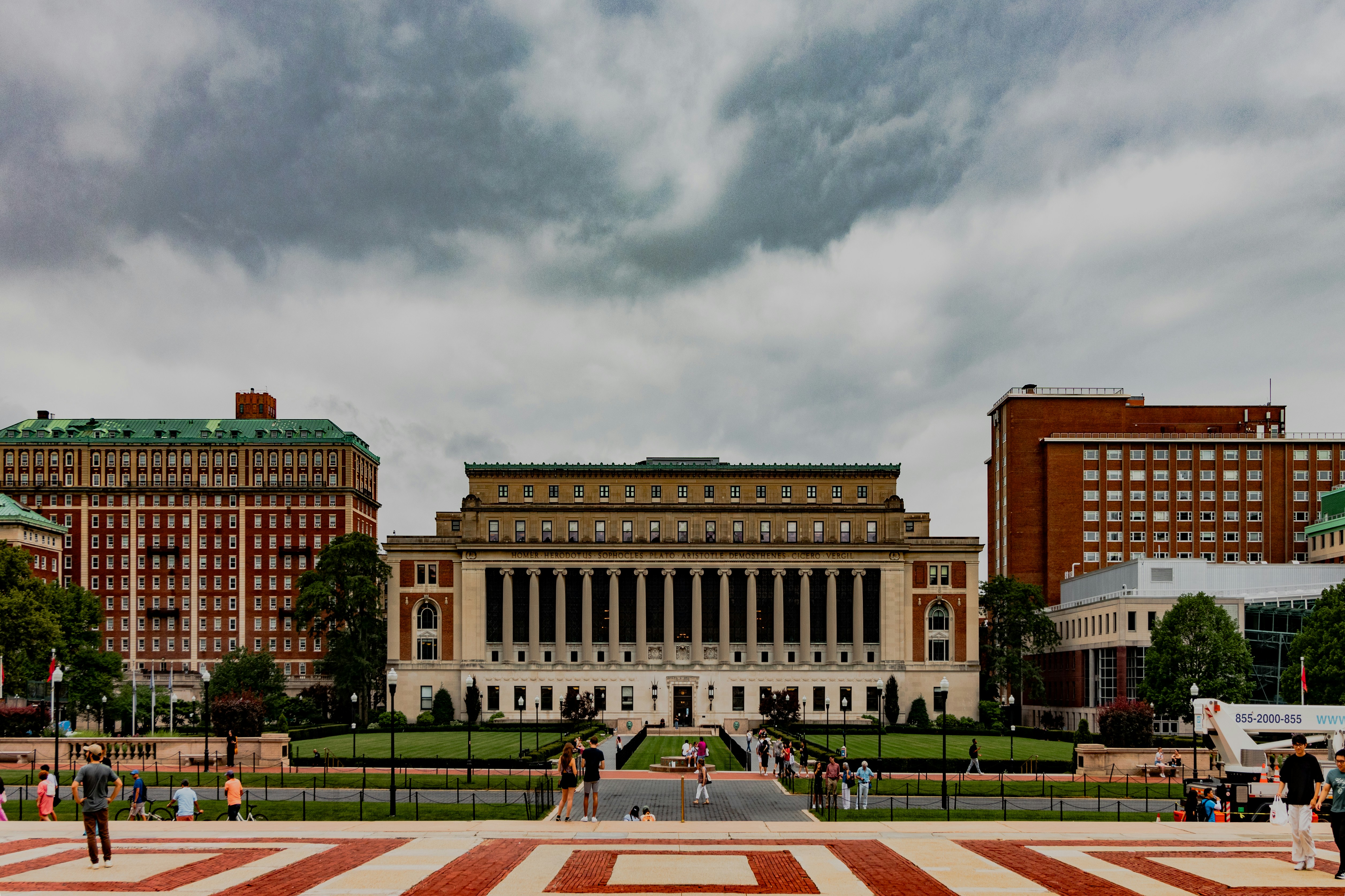 Neoclassical building with imposing columns stands against turbulent clouds, framed by greenery and red brick path.