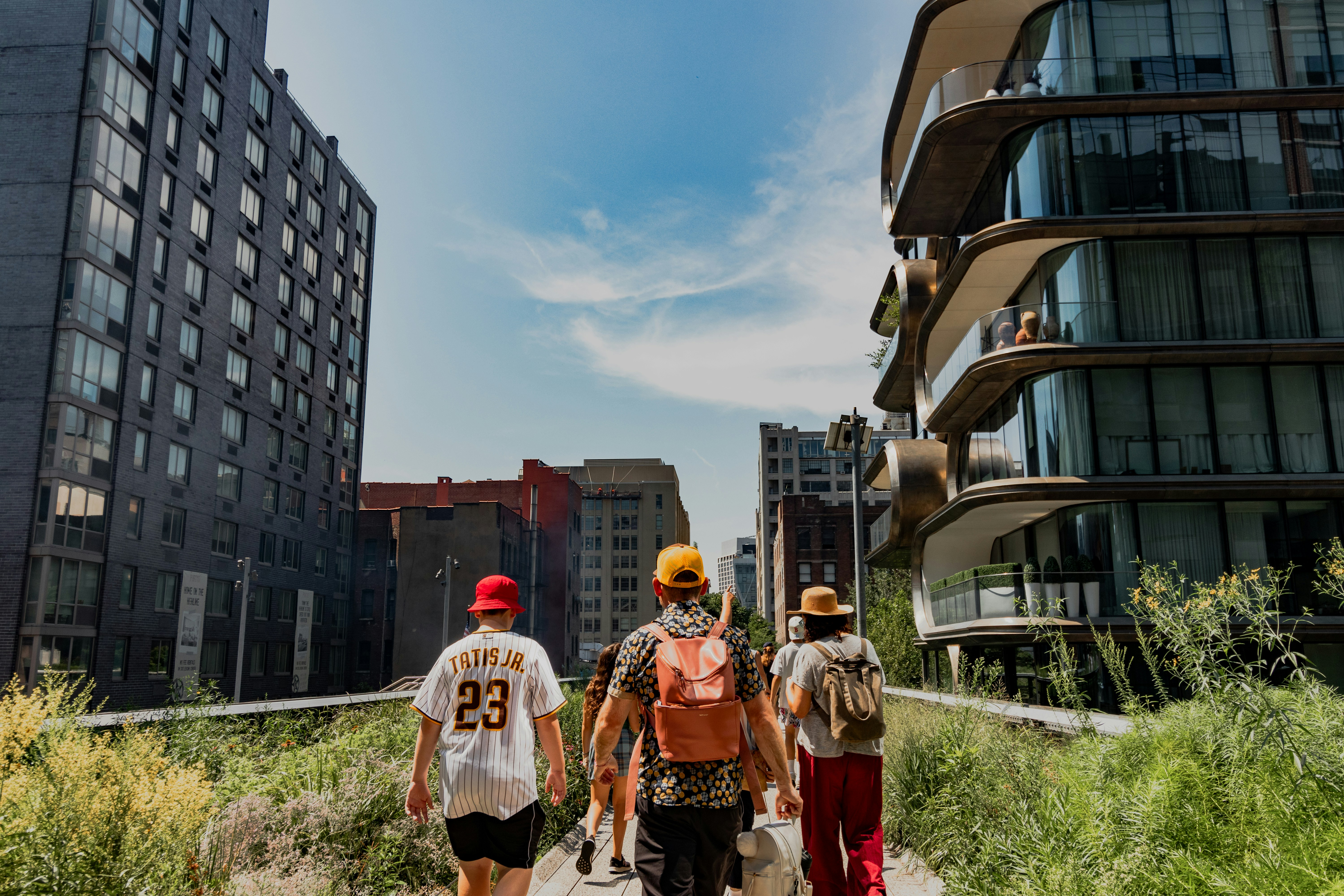 People walking on a lush elevated path with contrasting modern glass and traditional brick buildings under a clear blue sky.