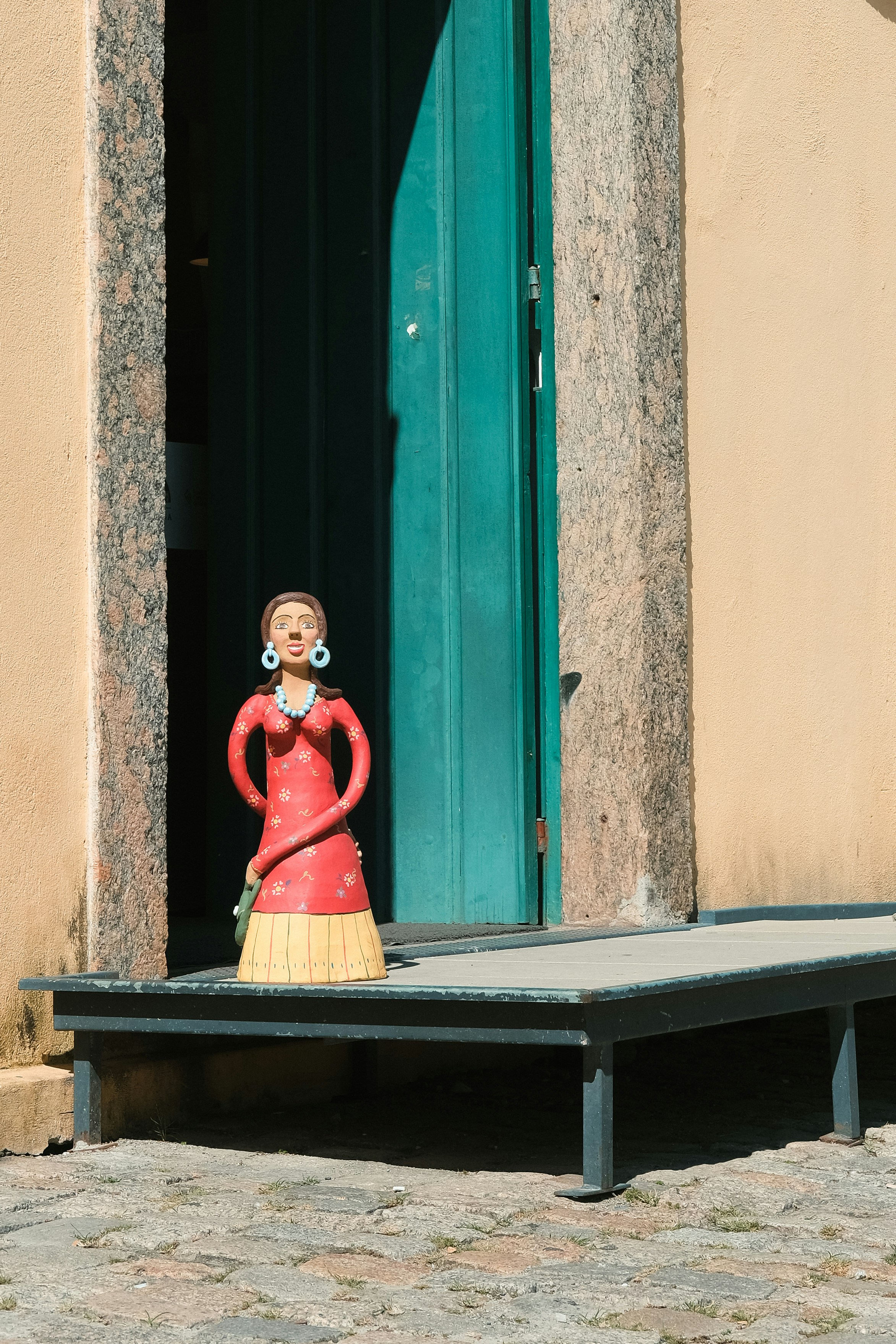 Red-Dressed Figurine Guarding a Teal Door on a Sunlit Porch