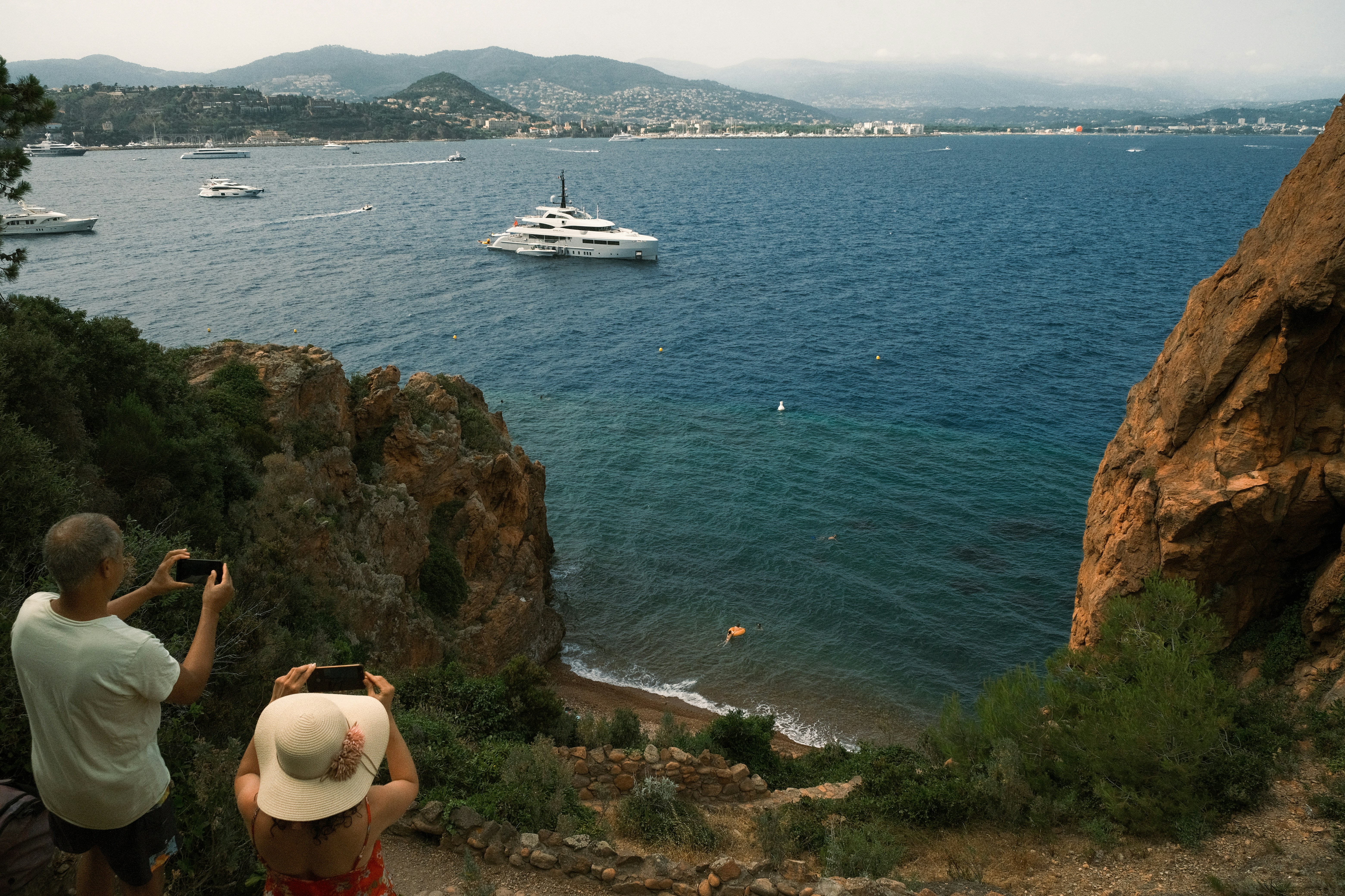 Yacht cruising along a vibrant blue sea framed by rocky cliffs and distant mountains under a hazy sky.