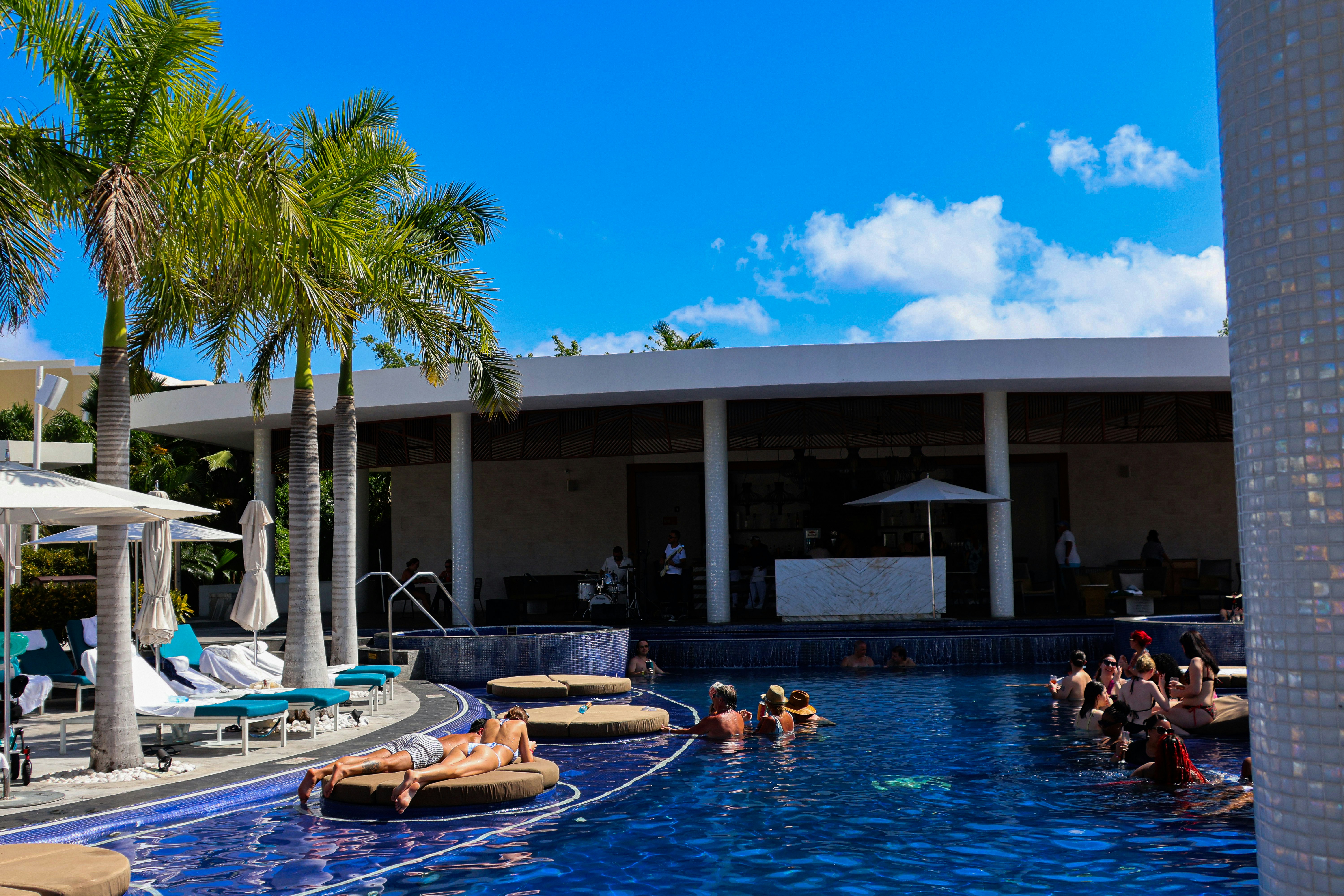 Palm trees sway over a vibrant outdoor pool filled with people enjoying a sunny day.